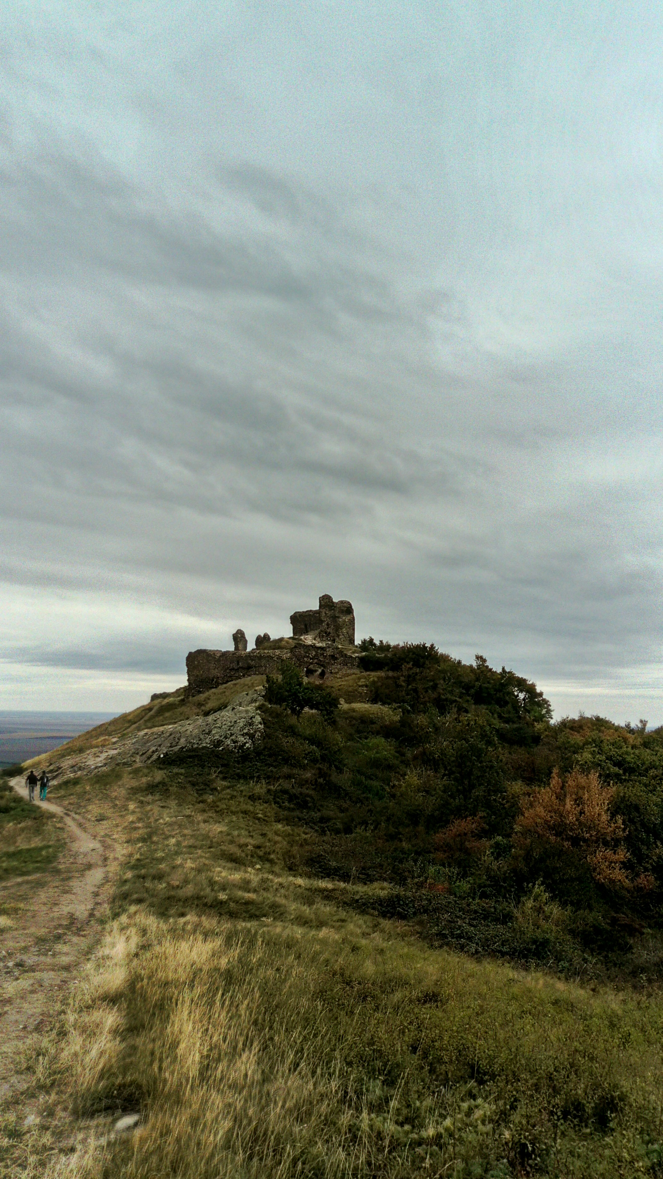 Ancient ruins perched atop a grassy hill, surrounded by a winding path and autumn foliage. The scene captures a moment of exploration against a moody sky.