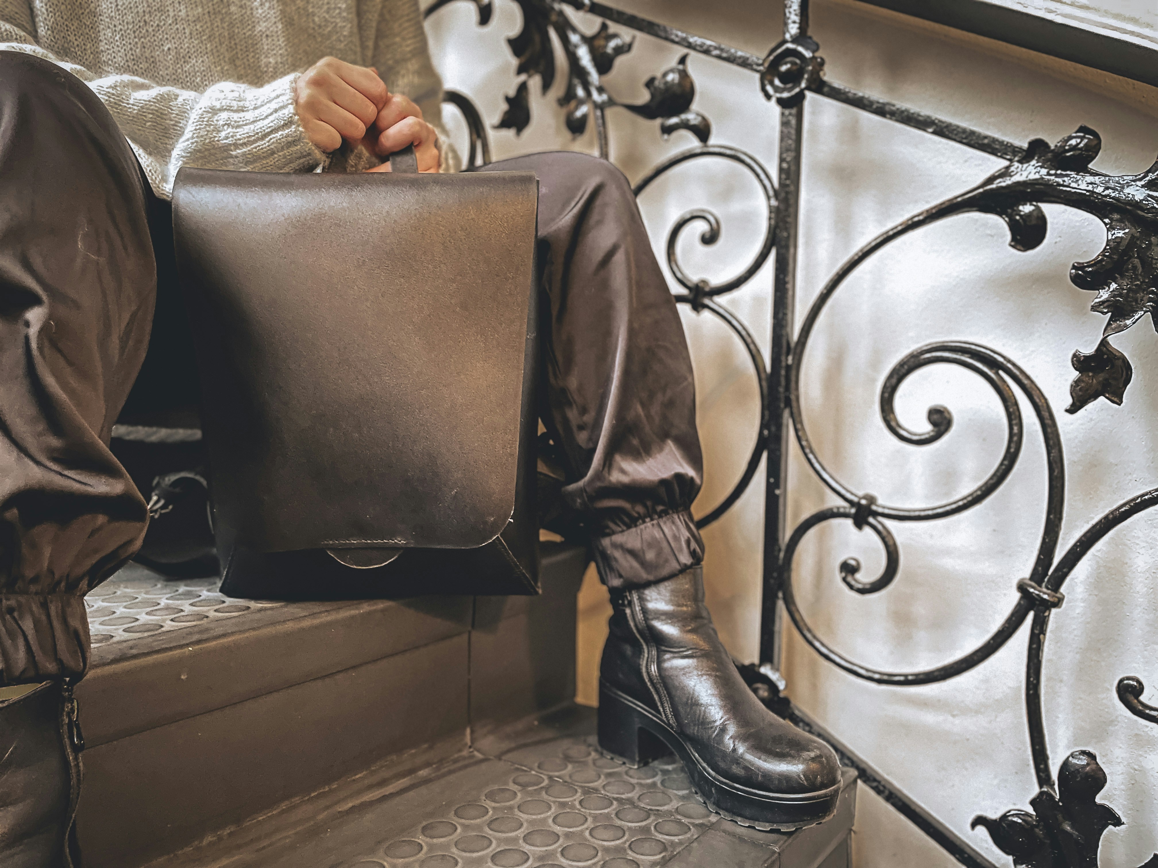 A person seated on a staircase, holding a sleek black bag, with intricate wrought iron railings in the background.