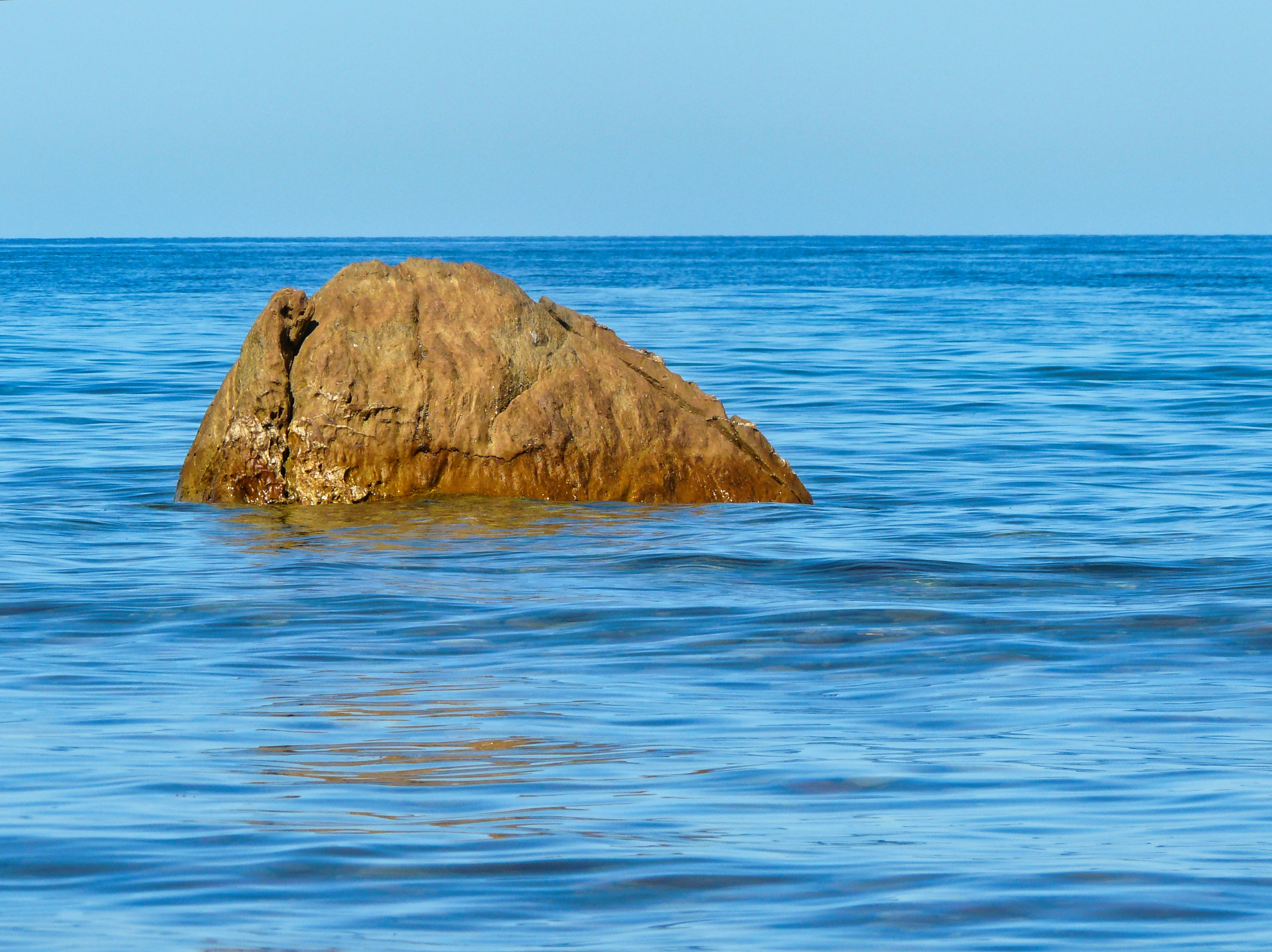 A rugged rock emerges from calm ocean waters under a clear blue sky, highlighting the tranquility of the coastal landscape.