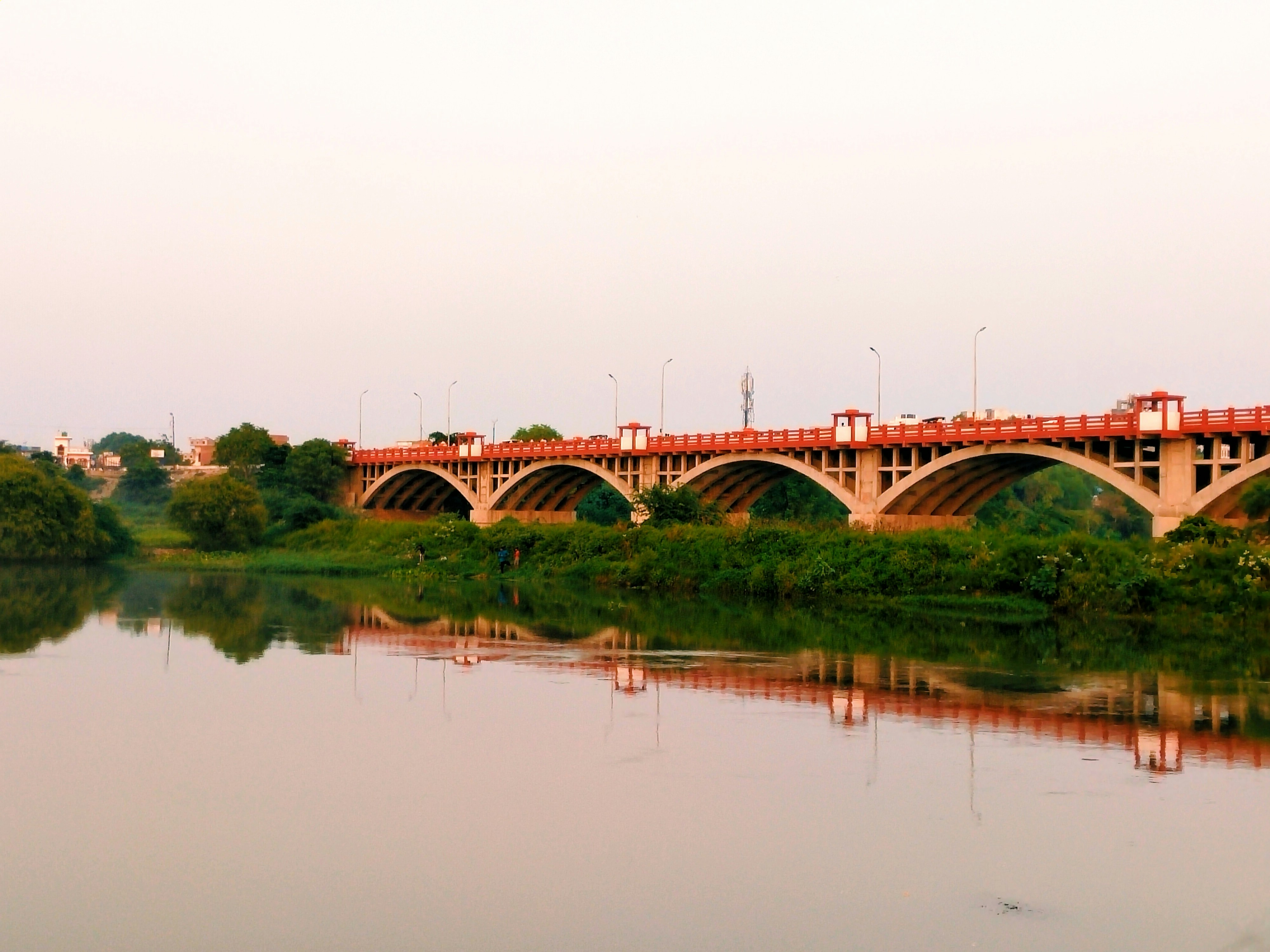 A red-arched bridge spans a calm river at dusk, its warm reflection shimmering on the water.