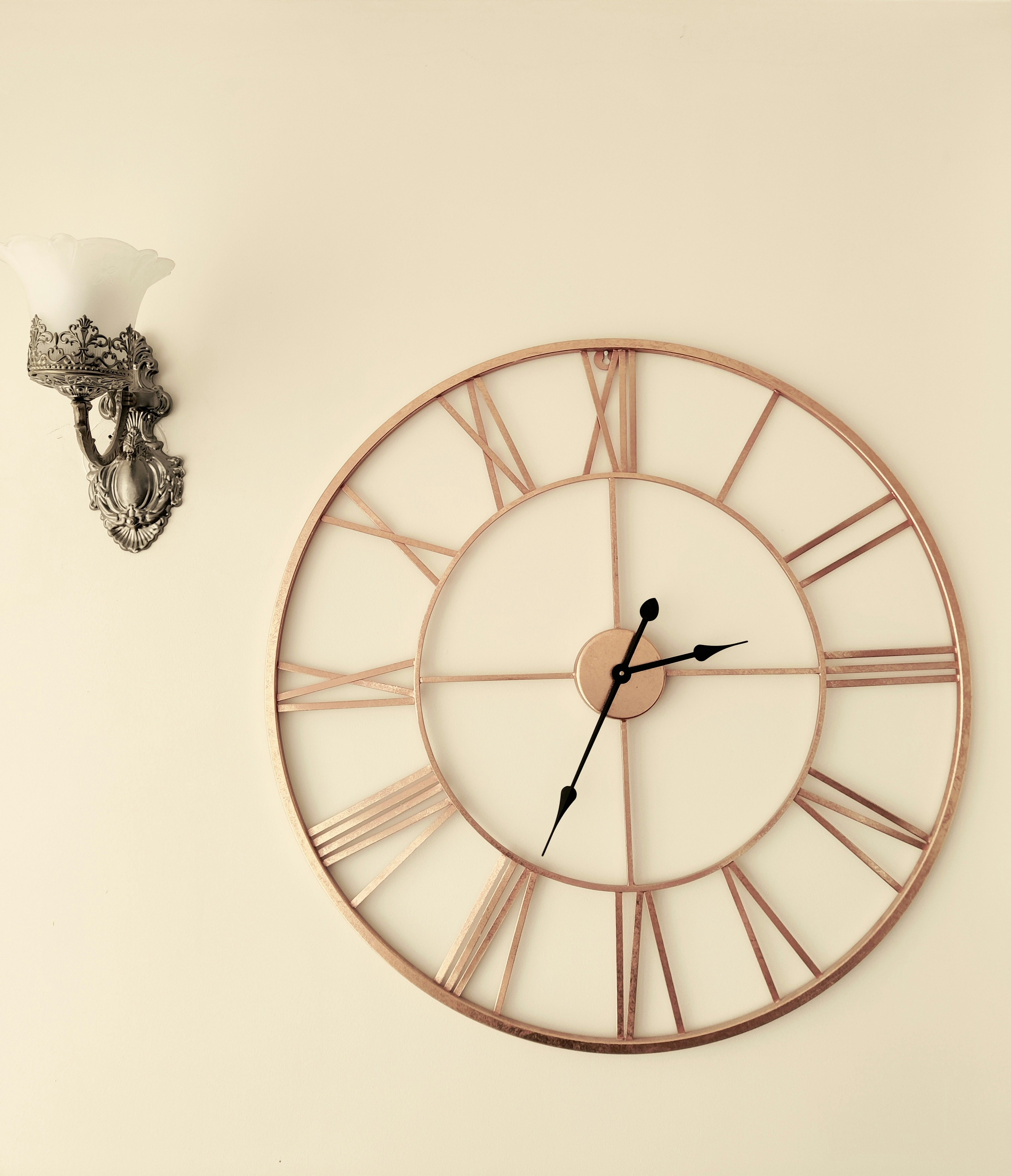 Copper-rimmed Roman numeral wall clock dominates a pale cream wall. An ornate antique sconce sits to the left, adding vintage detail.