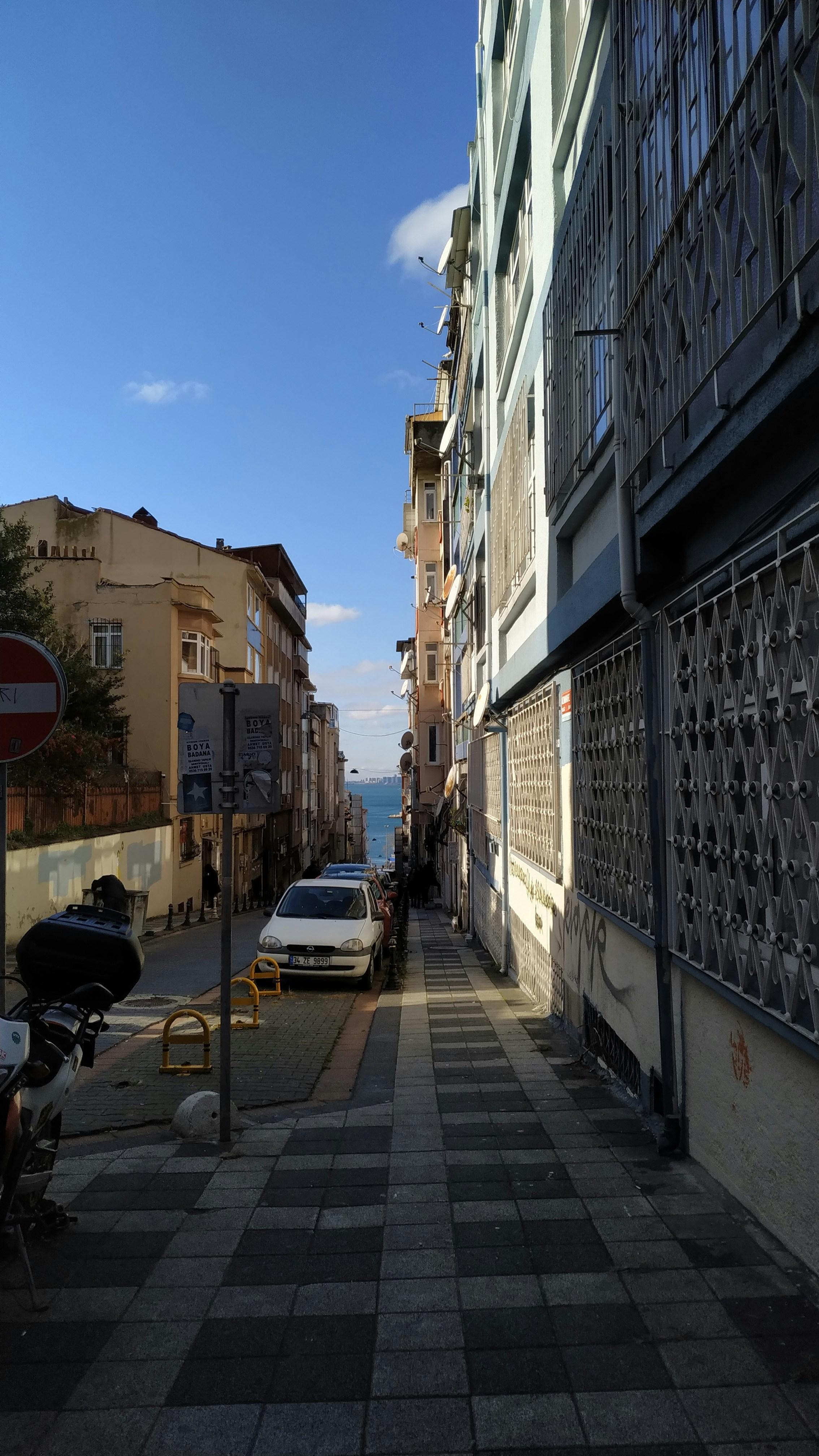 Narrow coastal alley flanked by pastel buildings and parked cars, converging toward a distant sea horizon.