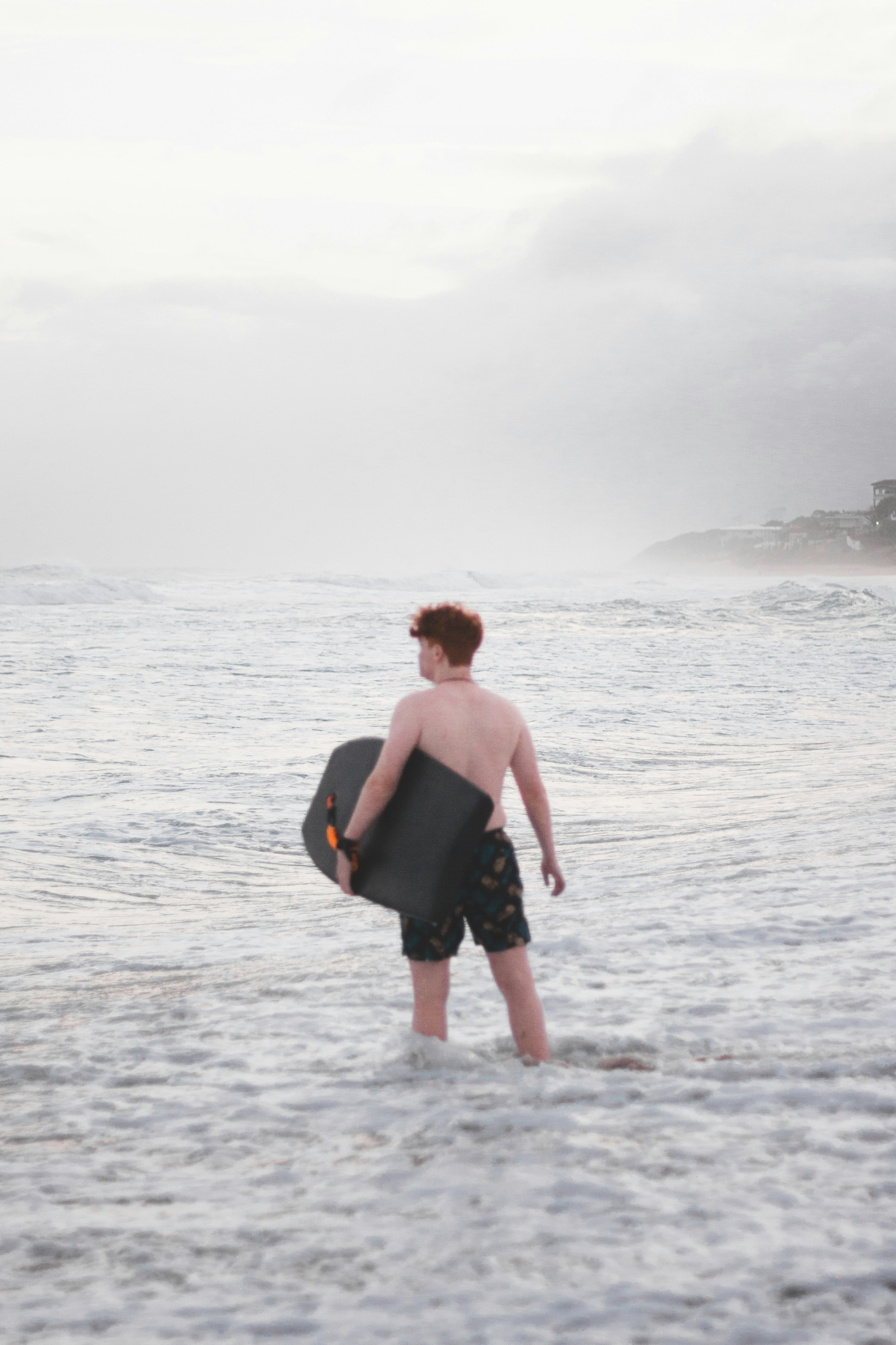 Lone bodyboarder steps into a misty sea, back to the viewer.