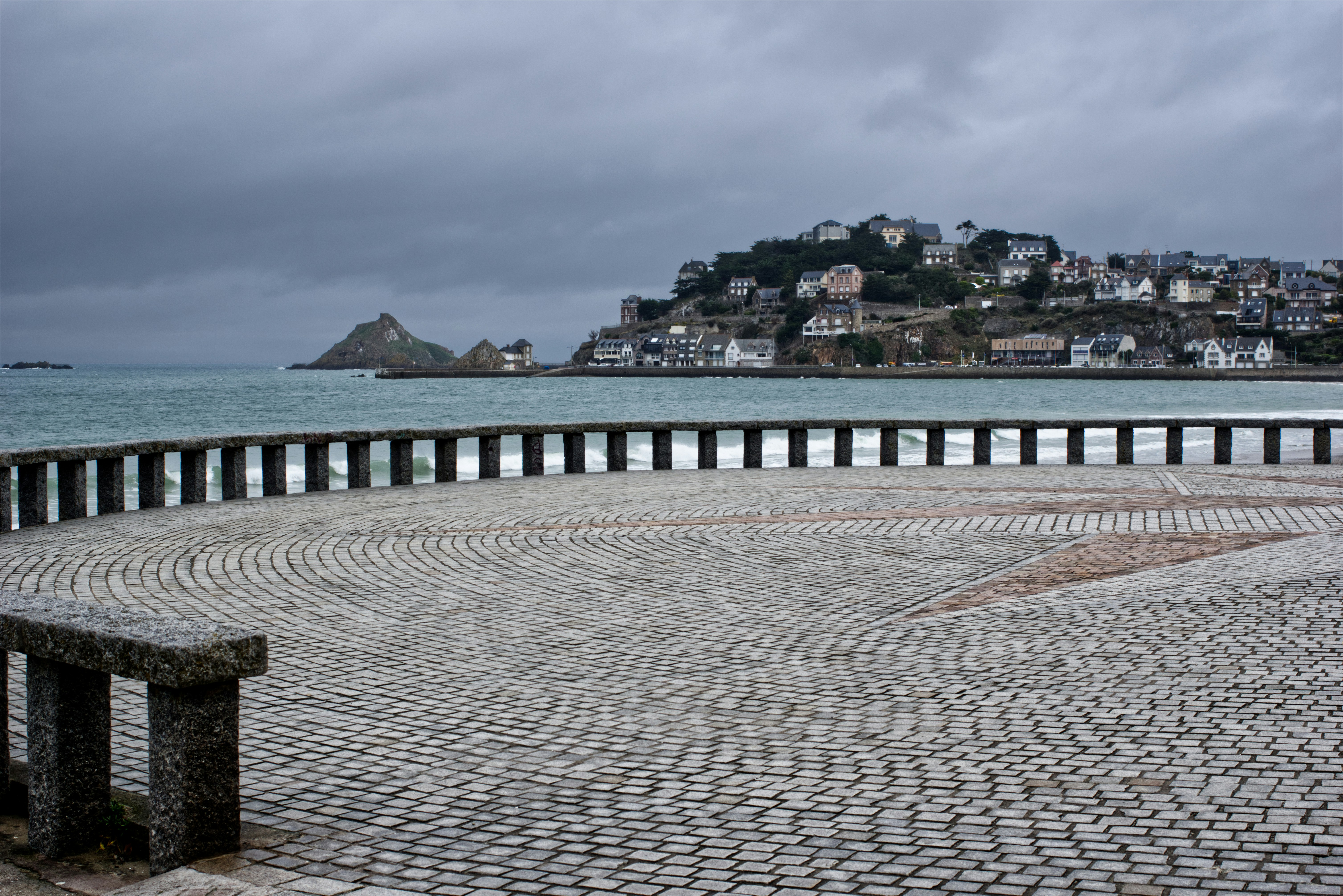 Circular stone promenade leading to a tranquil coastline, with a distant hillside village under an overcast sky.