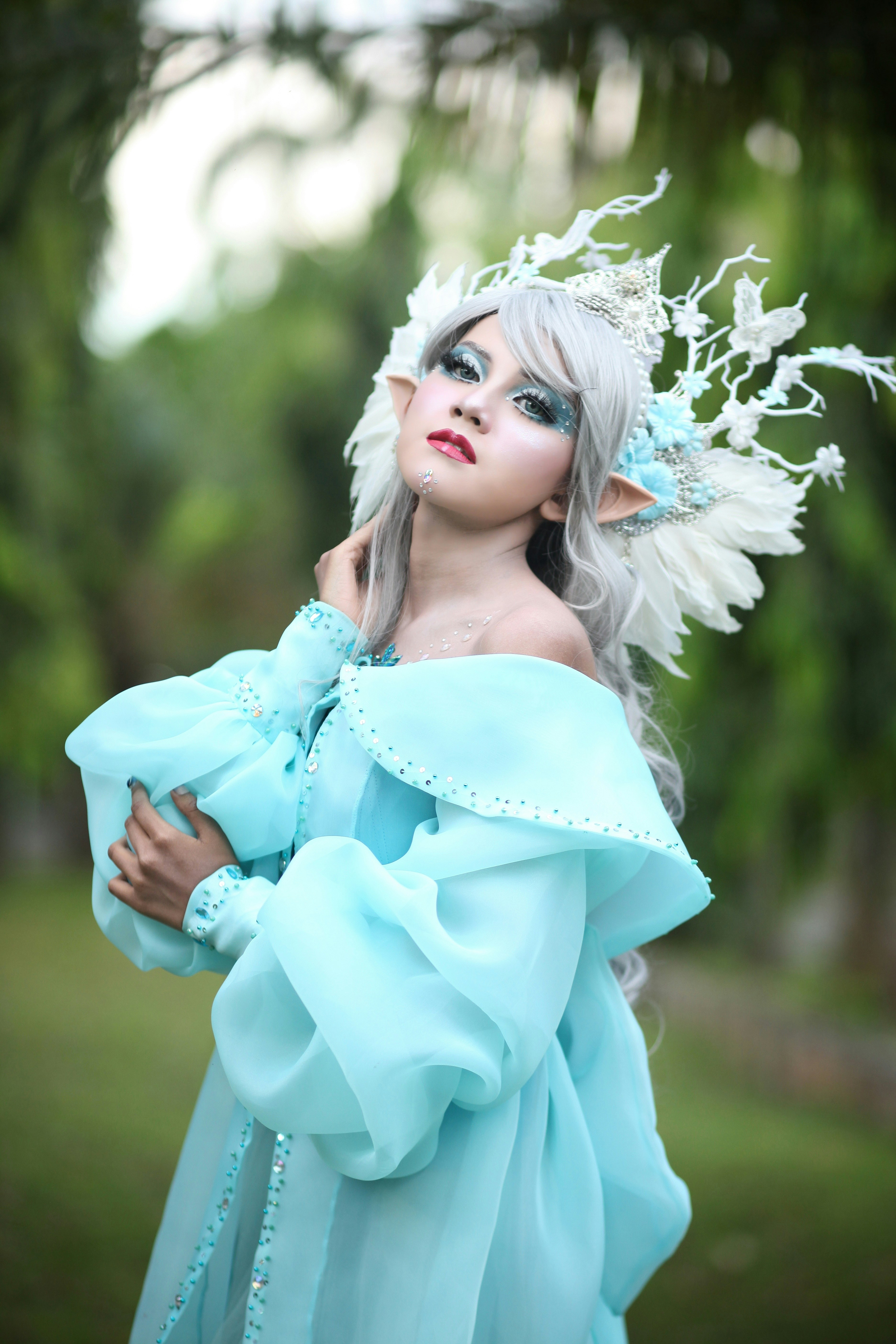 A character adorned in a flowing blue gown, embodying an ethereal winter spirit with delicate features and intricate accessories, set against a lush green backdrop.