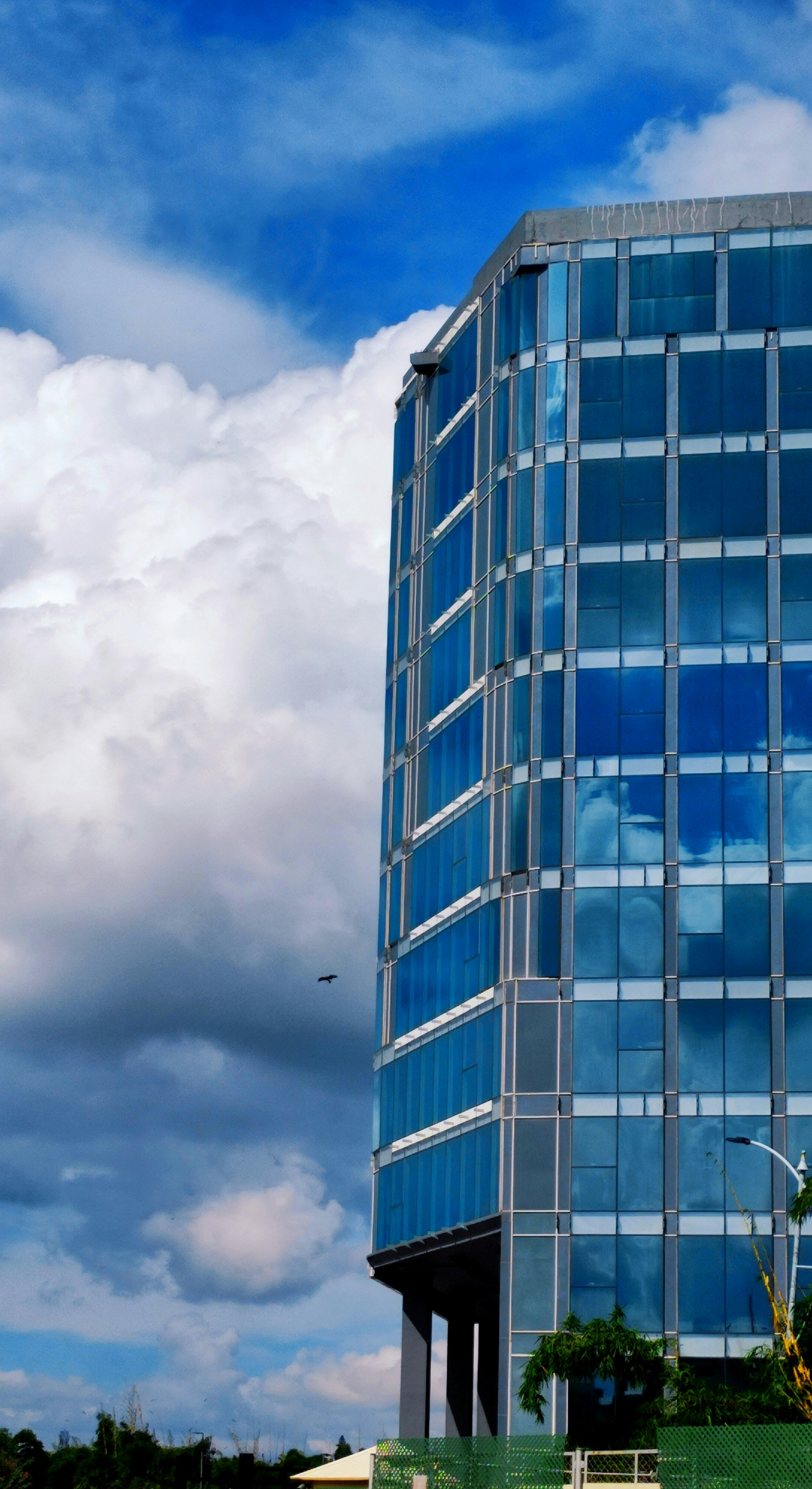 A glass-clad office tower rises along the right edge against a vivid blue sky and billowy clouds. The scene highlights the building's grid of reflective windows and clean architectural lines.
