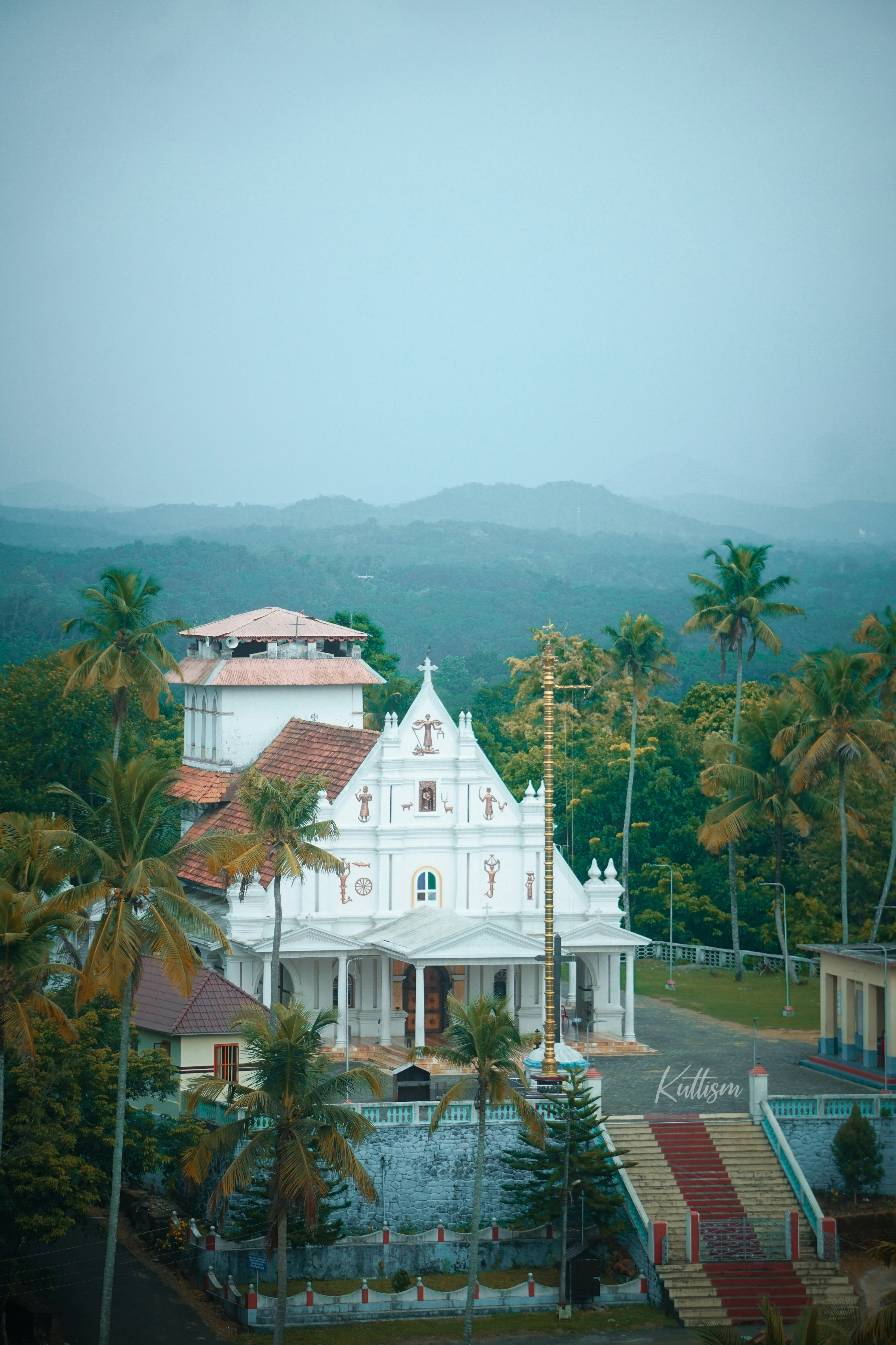 A majestic white church stands amidst lush greenery, framed by distant mountains under a soft, overcast sky.
