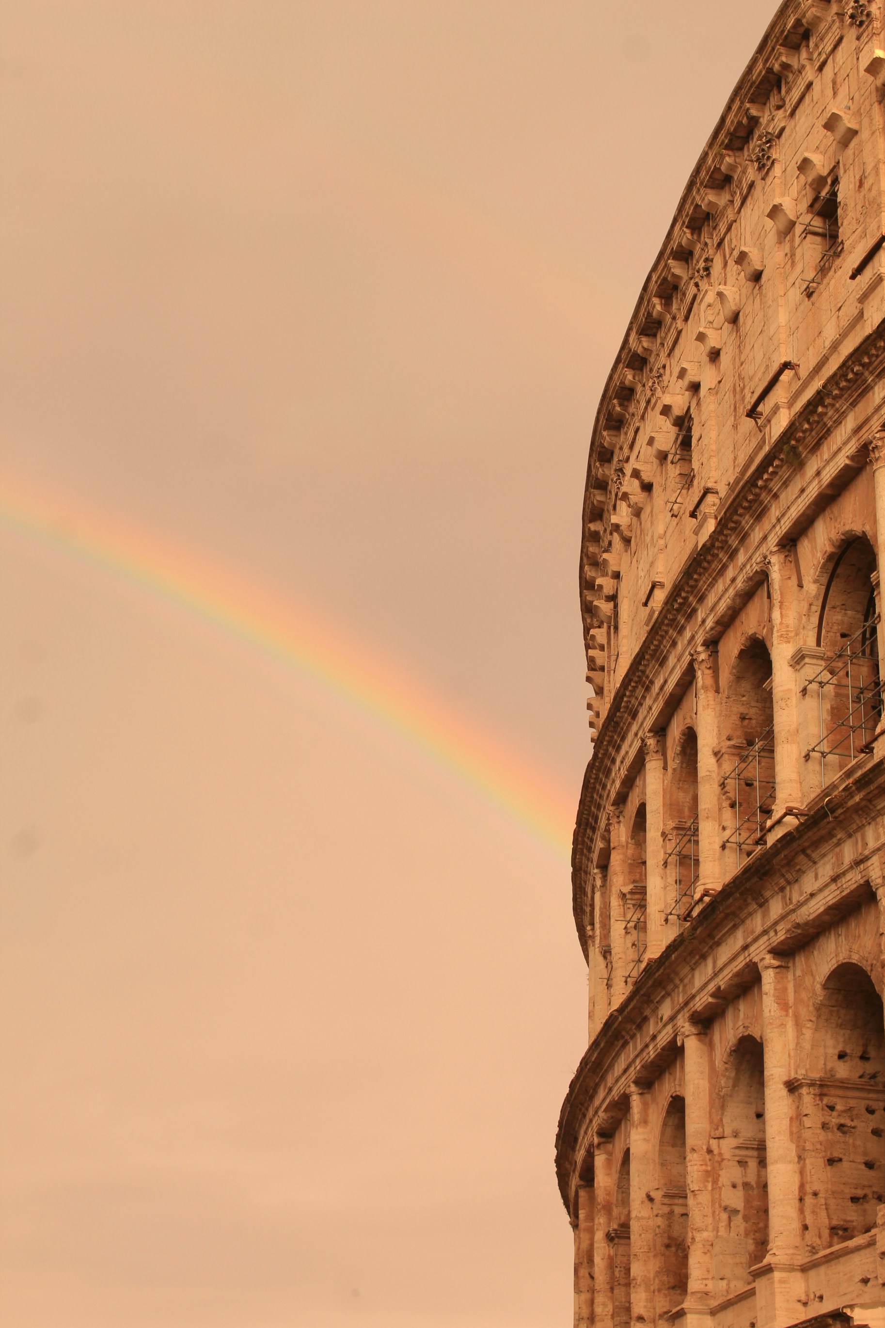 The Colosseum stands majestically against a pastel sky, framed by a delicate rainbow arching above. The scene captures the timeless beauty of Rome.