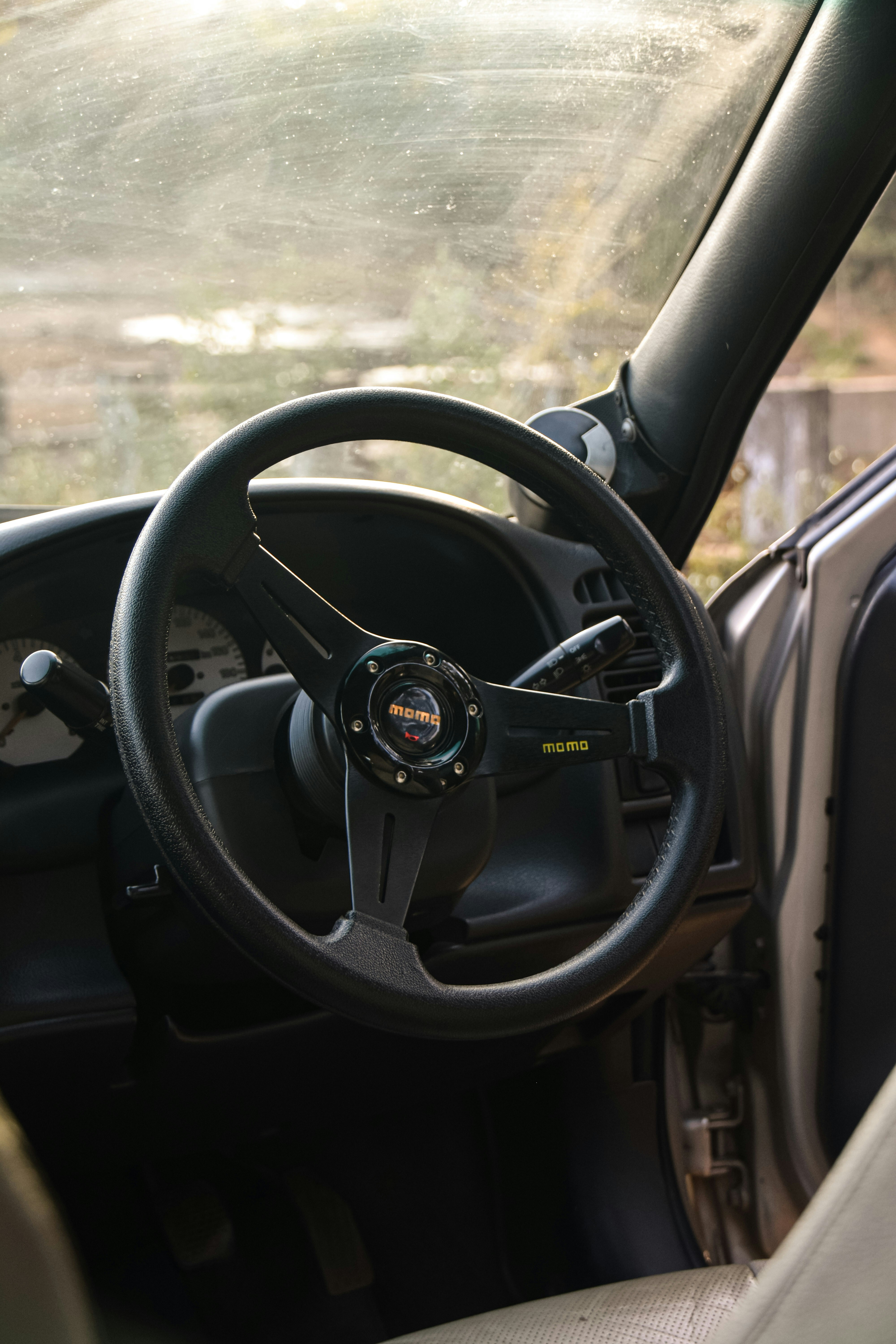Close-up of a Momo steering wheel inside a vehicle, showcasing its sleek design and the dashboard in soft focus. The image emphasizes the connection between driver and machine.