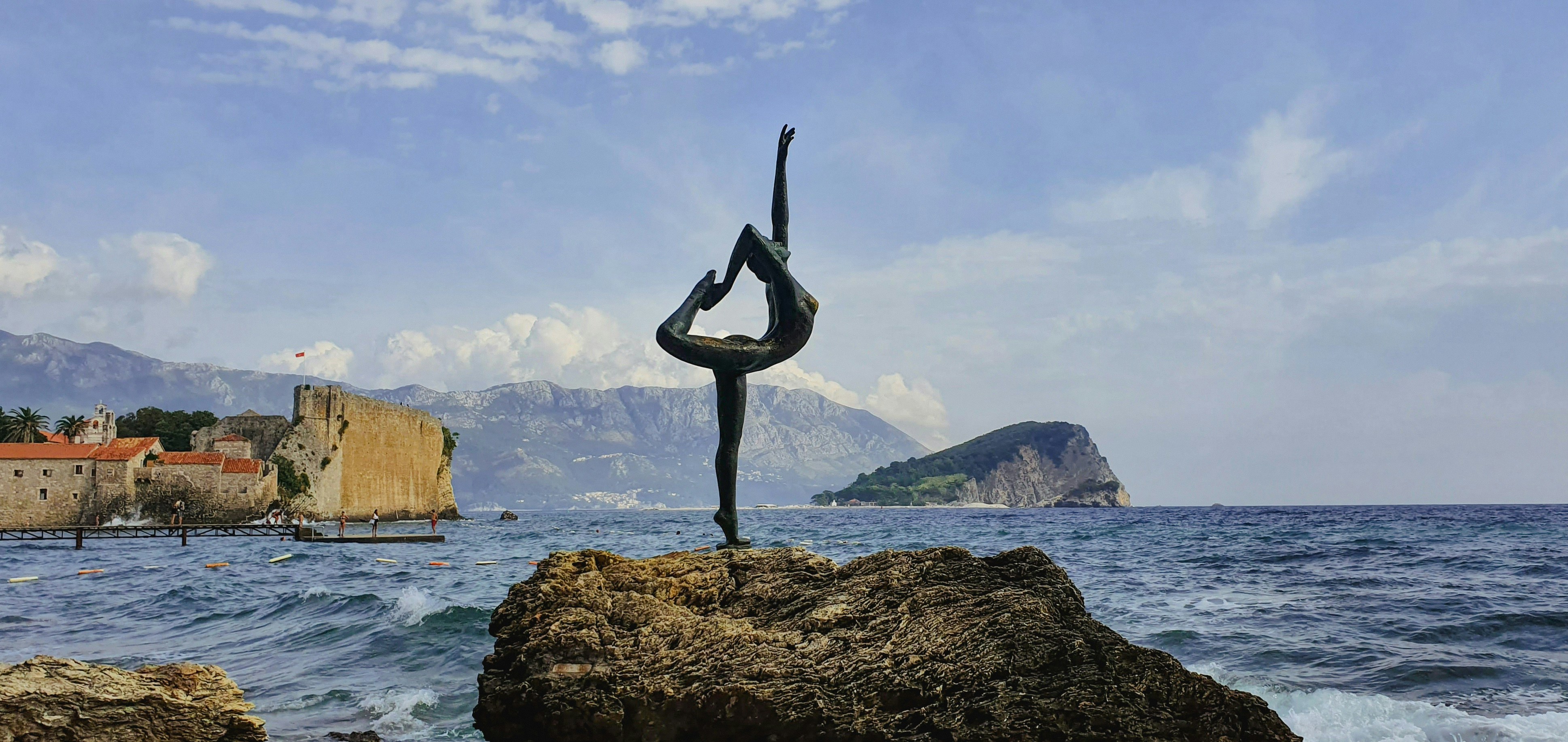 Bronze sculpture of a dancer poised gracefully on a rock by the sea, with mountains and a historic fortress in the background.