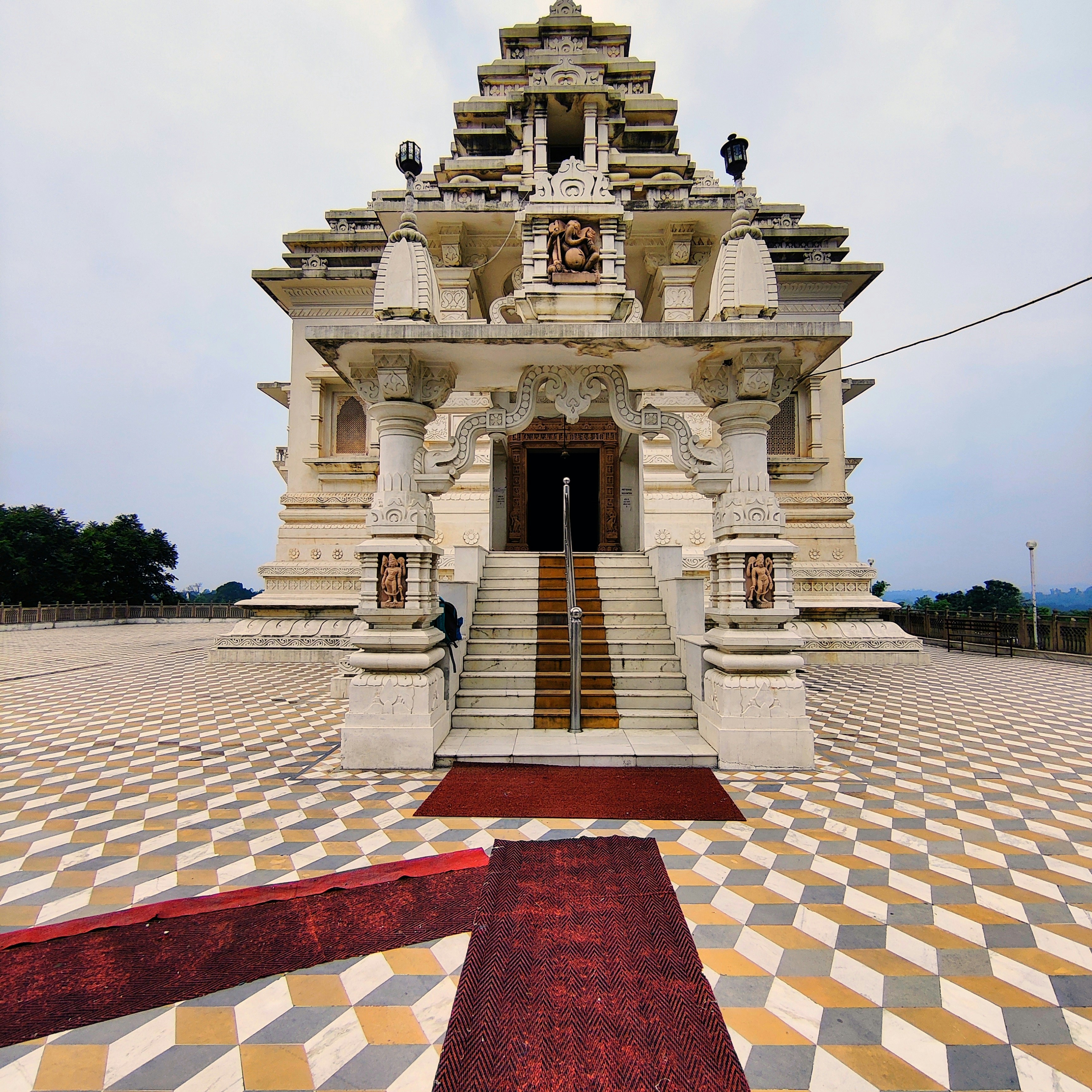 Ornate stone temple rises from a tessellated plaza. A red carpet leads up to its carved steps.