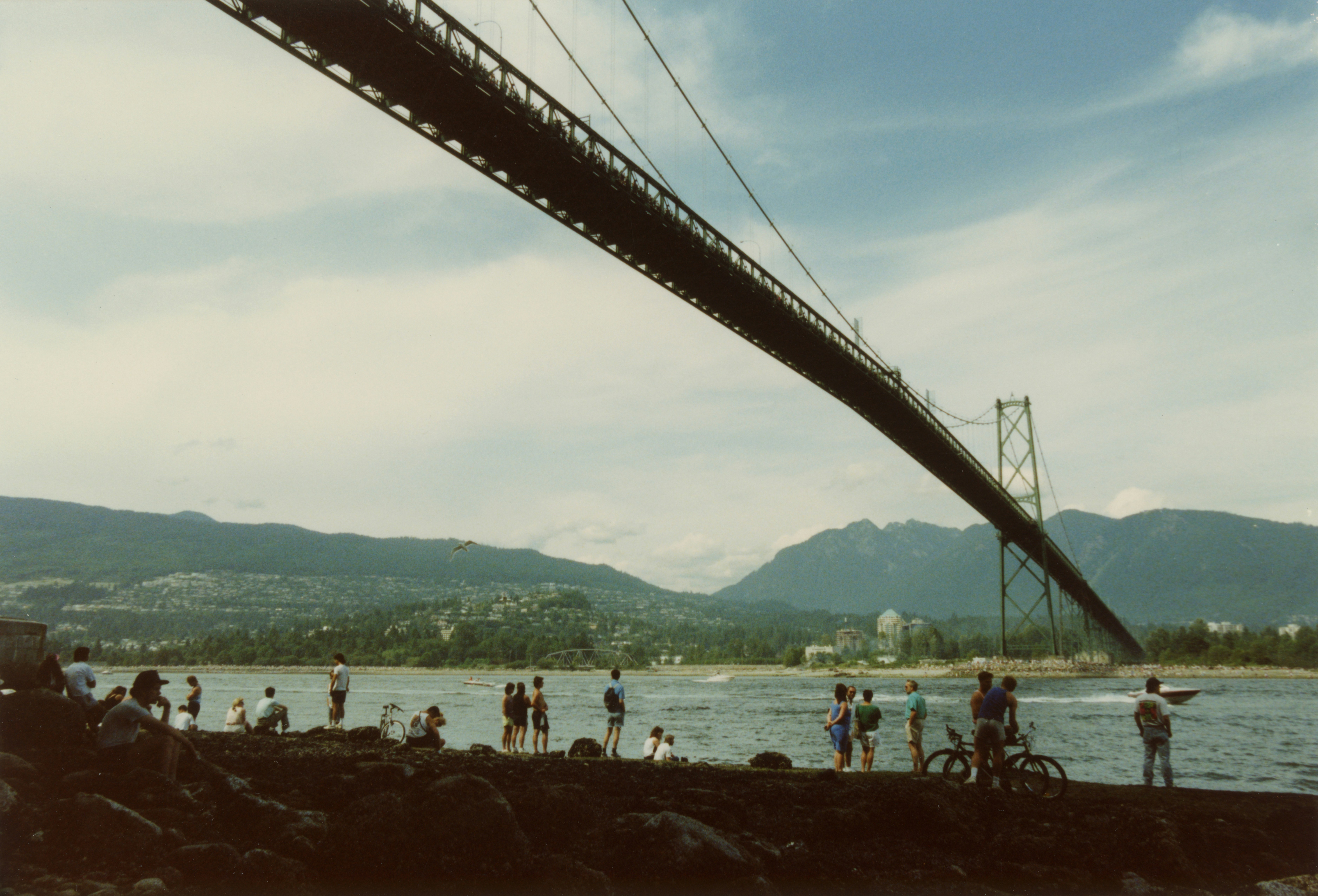 A photograph capturing a long suspension bridge spanning a calm bay, with people gathered on the rocky shoreline in the foreground.