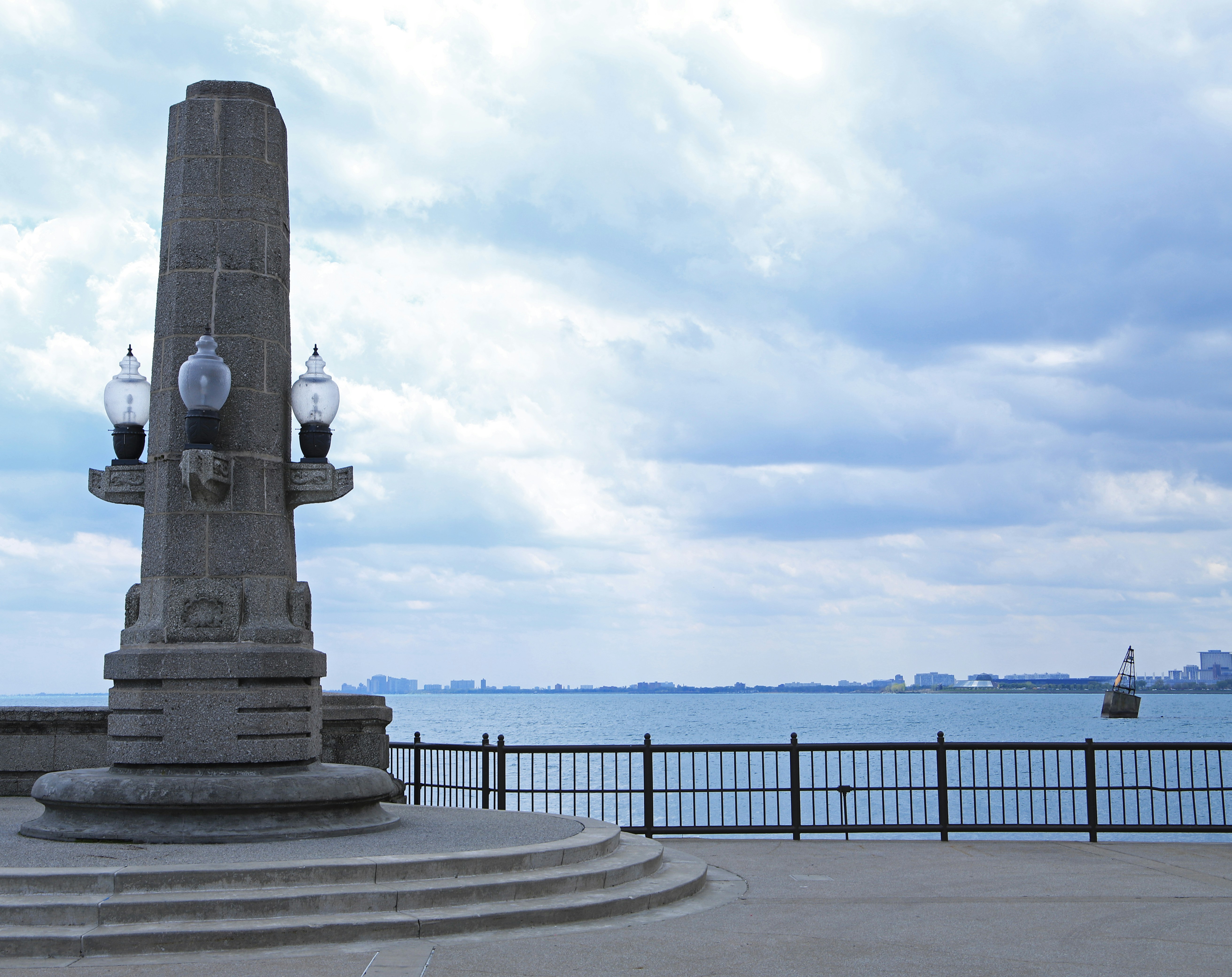 Historic stone monument with decorative lamps beside a calm lake, framed by a cloudy sky. The scene evokes a sense of tranquility and nostalgia.