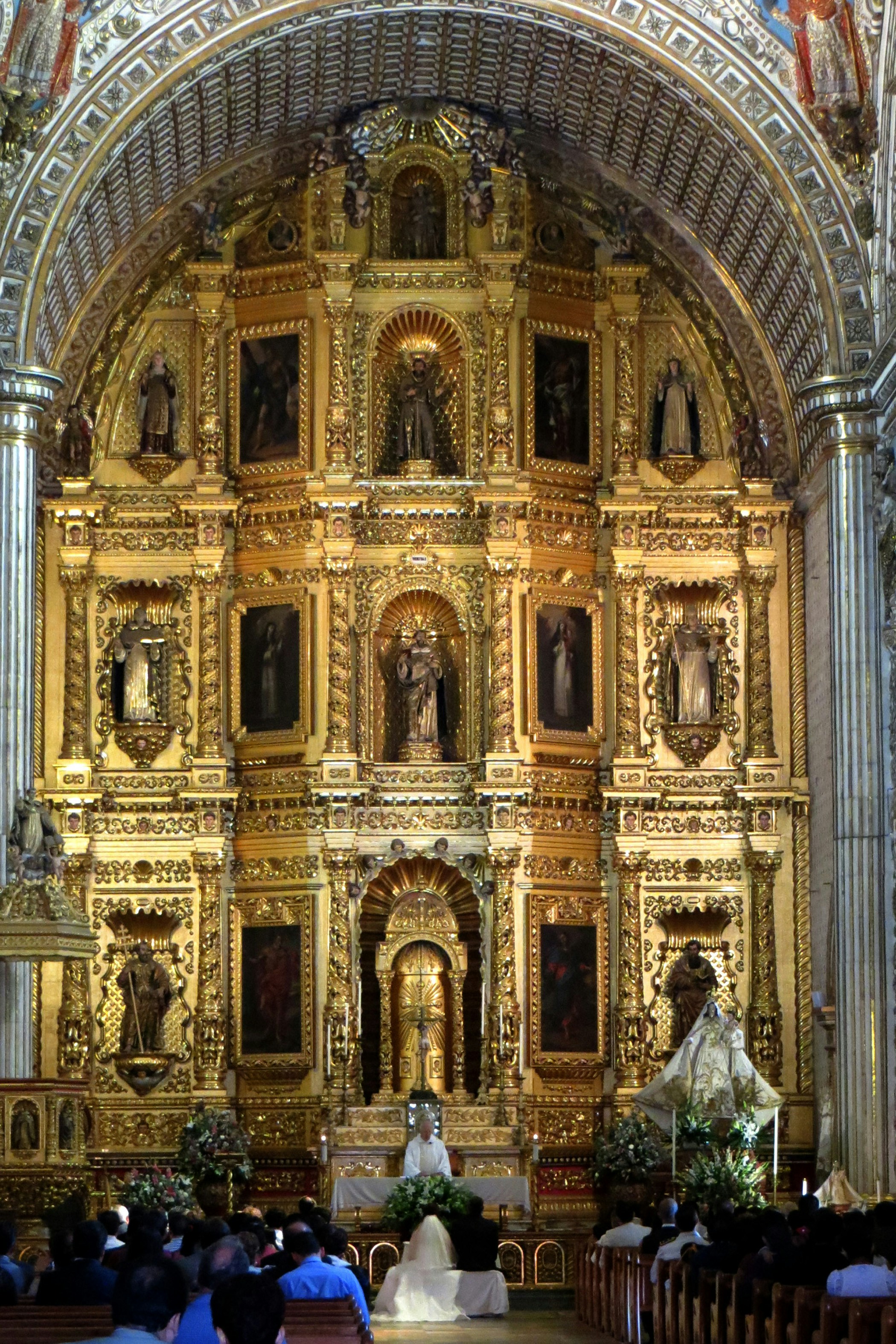 Intricate altar adorned with gold leaf and religious statues, capturing a solemn ceremony with attendees seated in reverence.