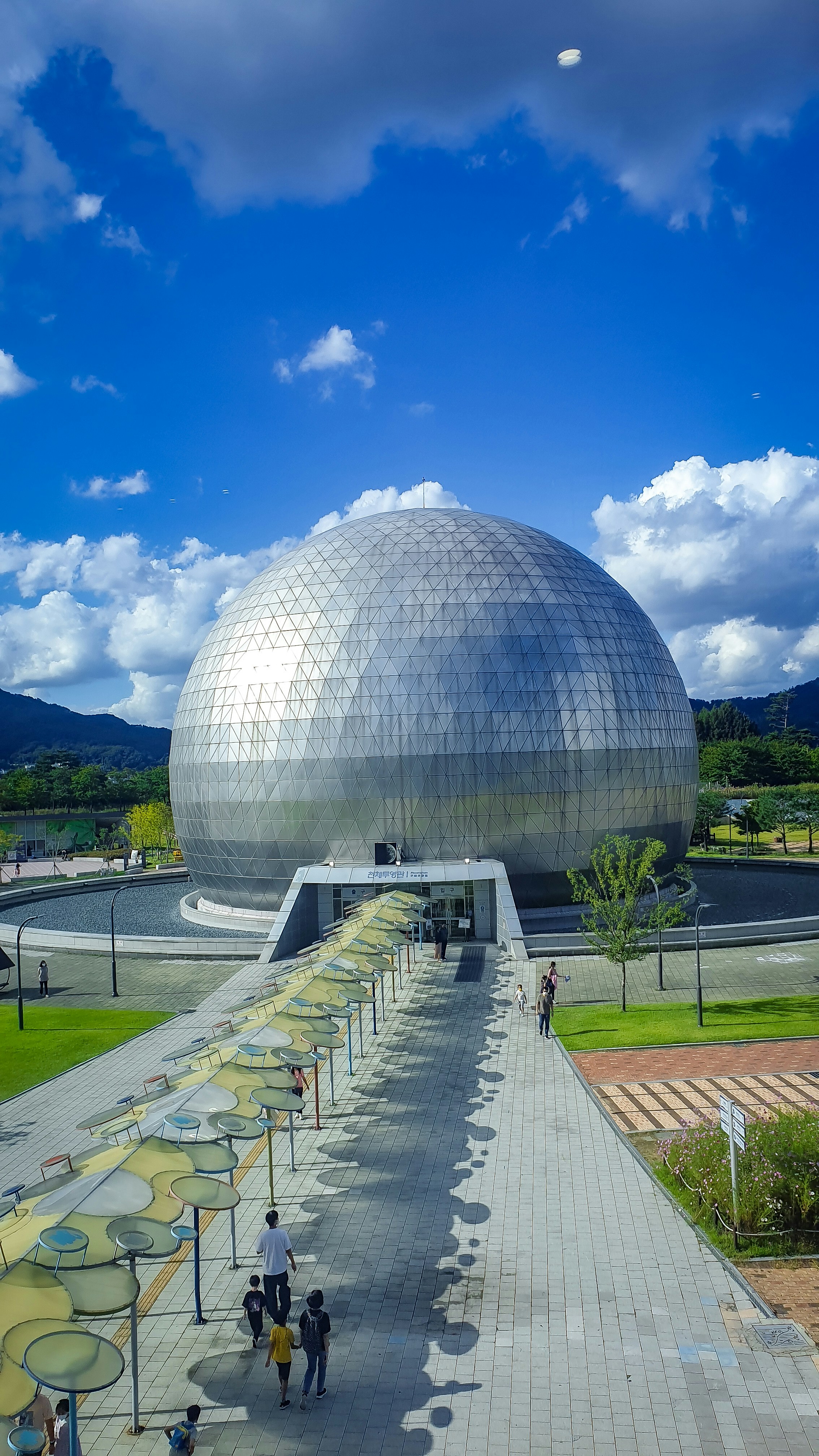 A large, reflective spherical building surrounded by greenery and a pathway, under a vibrant sky dotted with clouds.