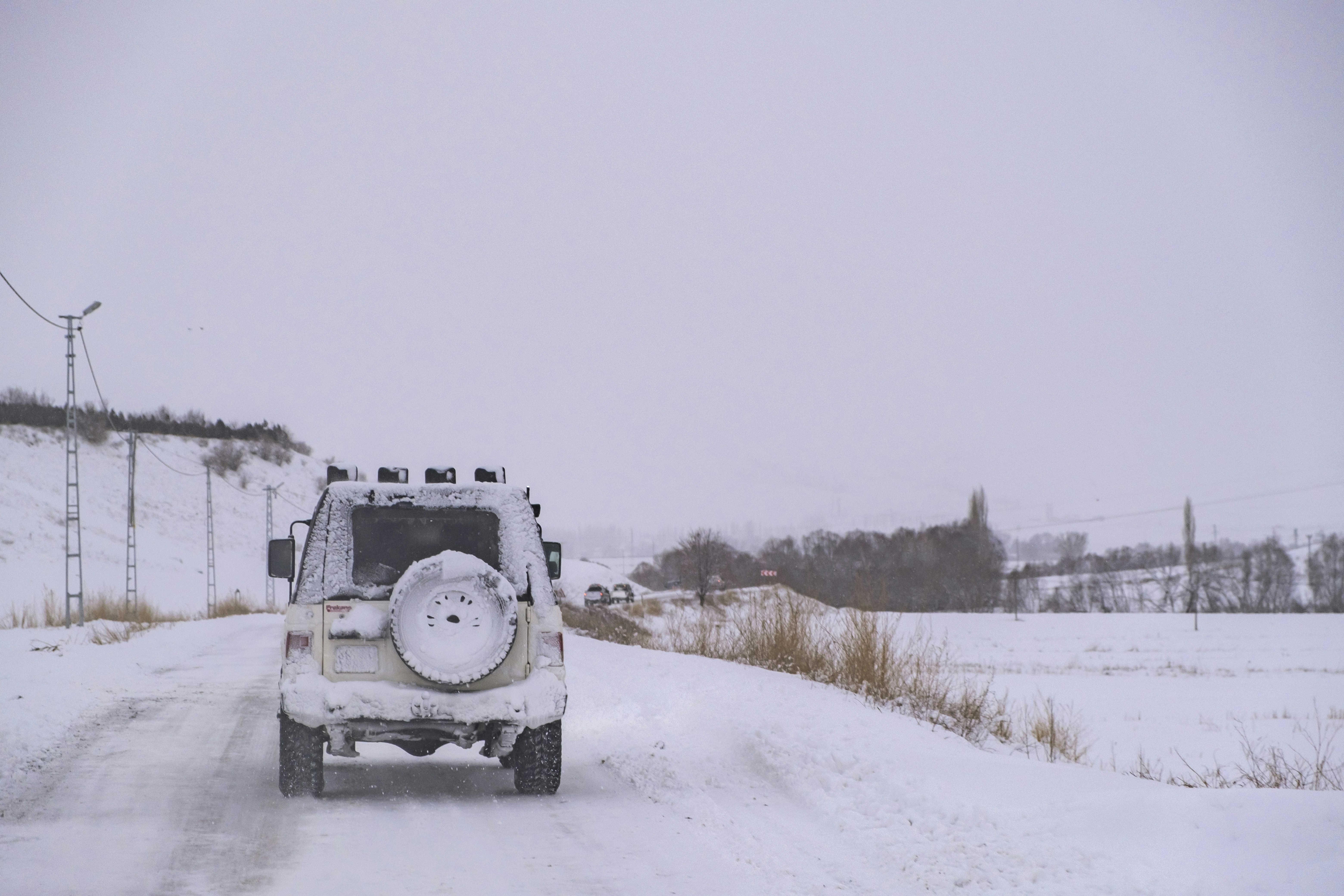 Off-road vehicle driving along a snow-covered rural road under a cloudy sky.