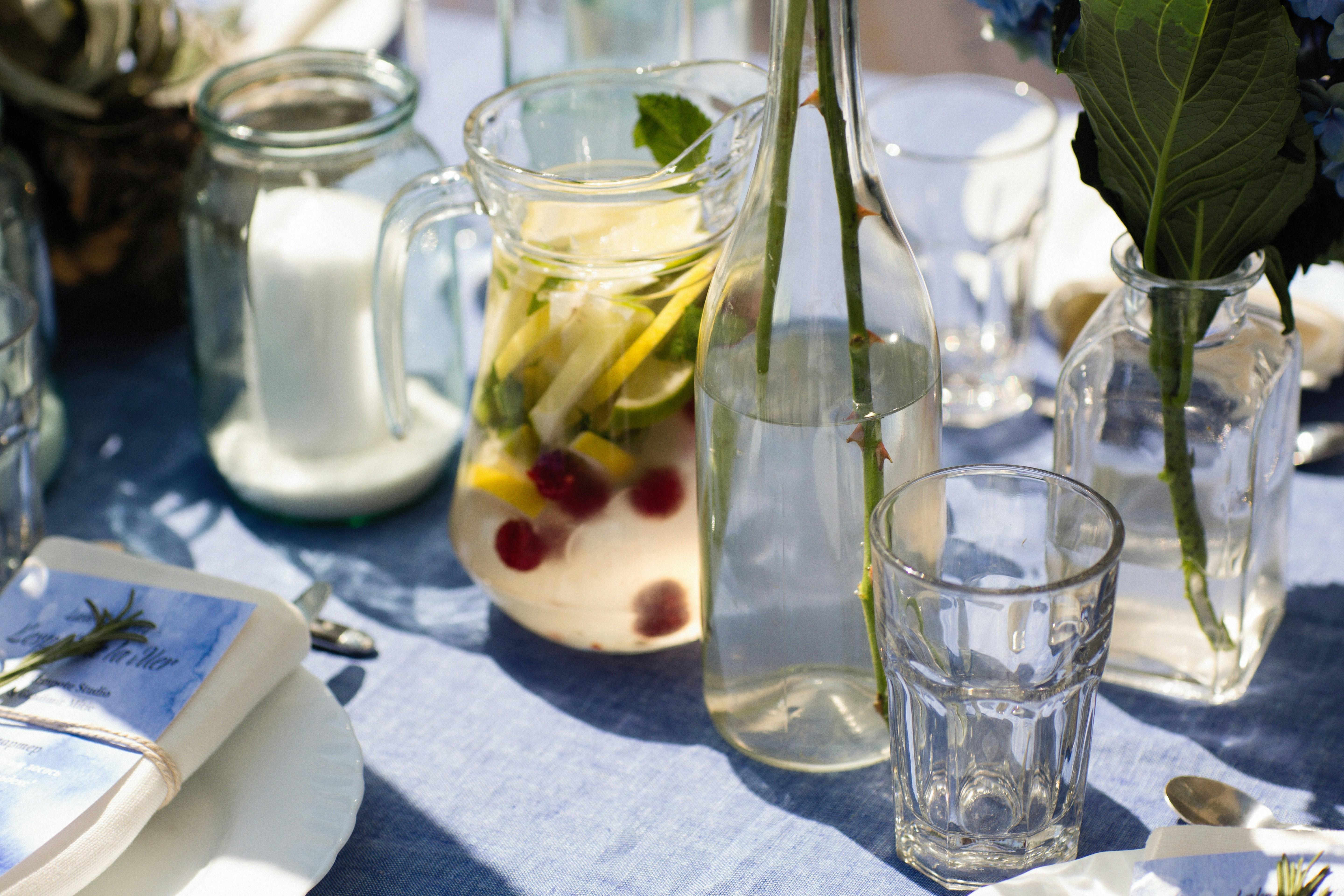 Table setting featuring a glass pitcher filled with fruits and herbs, surrounded by elegant glassware and floral arrangements. Perfect for a summer gathering.