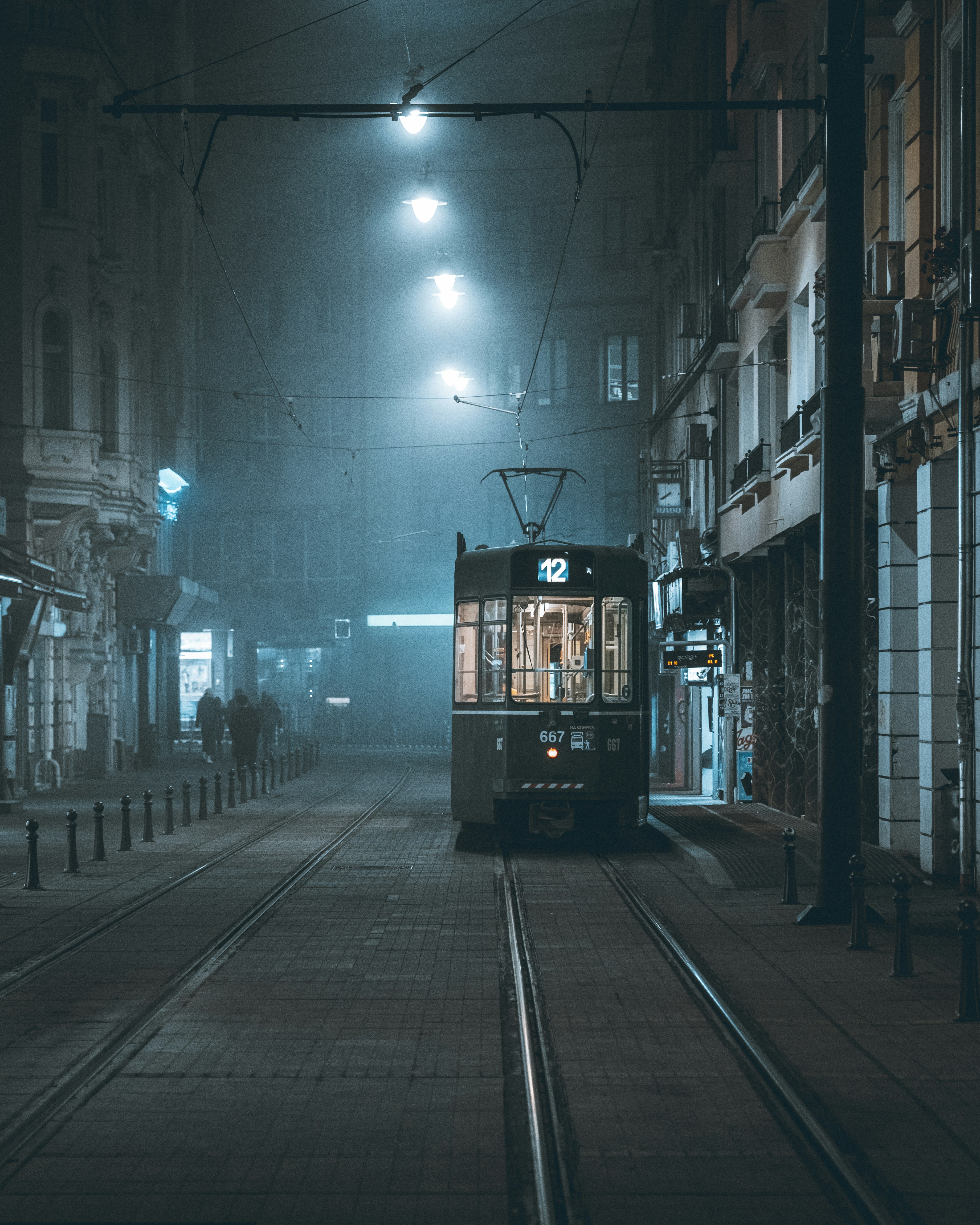 A vintage tram stands illuminated in a foggy urban street, flanked by dimly lit buildings and faint silhouettes of pedestrians. The atmosphere evokes a sense of nostalgia and mystery.