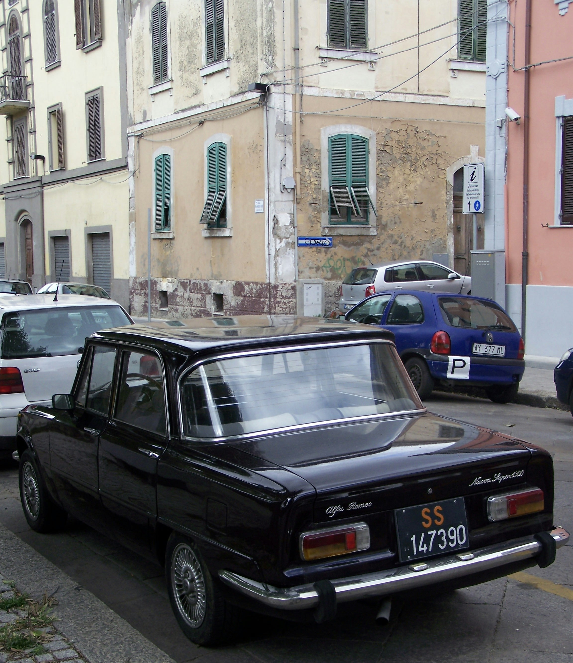 Classic Alfa Romeo parked on a quaint street with weathered buildings and colorful cars in the background.