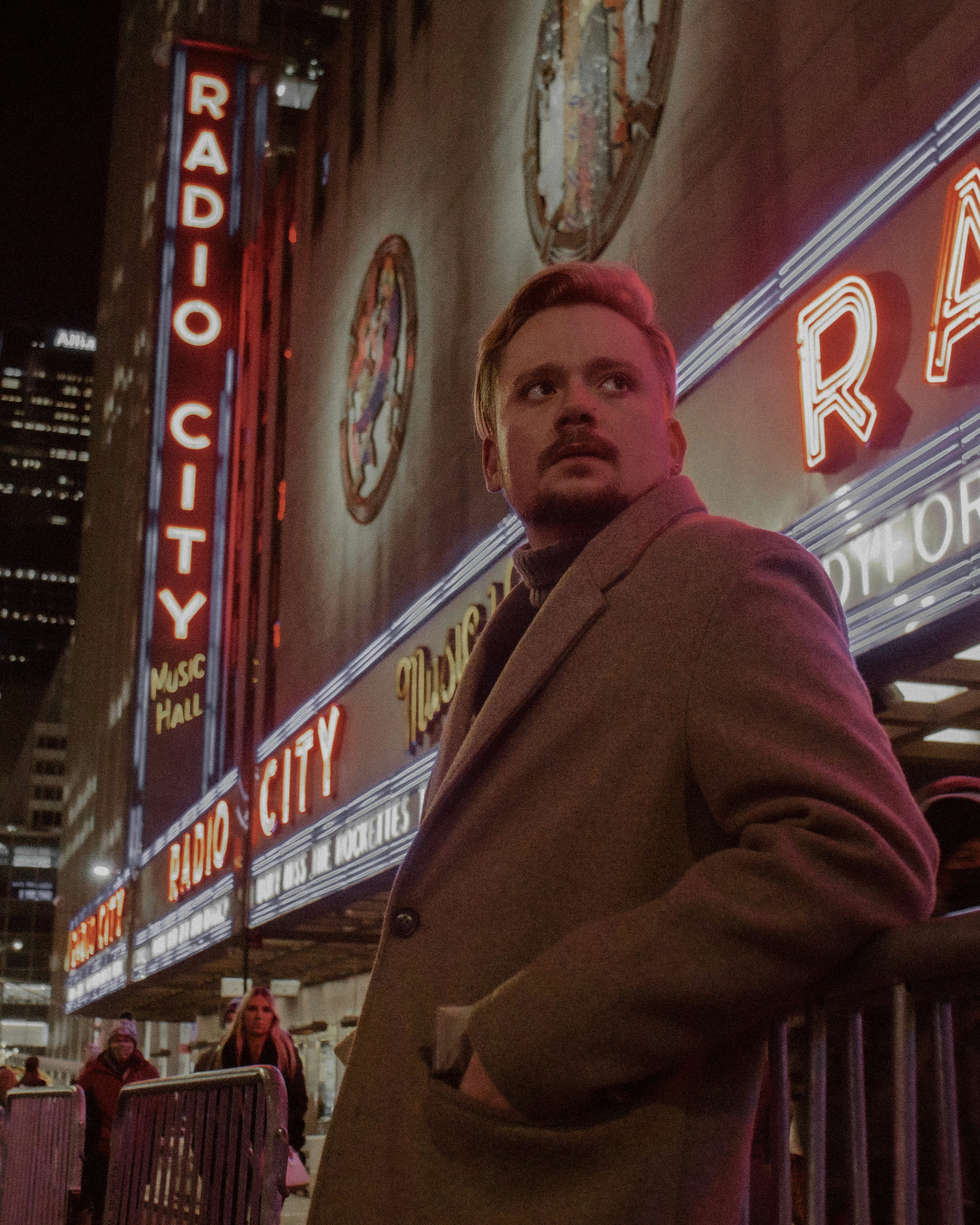 A man in a coat stands pensively outside Radio City Music Hall, illuminated by vibrant neon lights. The bustling city backdrop enhances the scene's urban atmosphere.