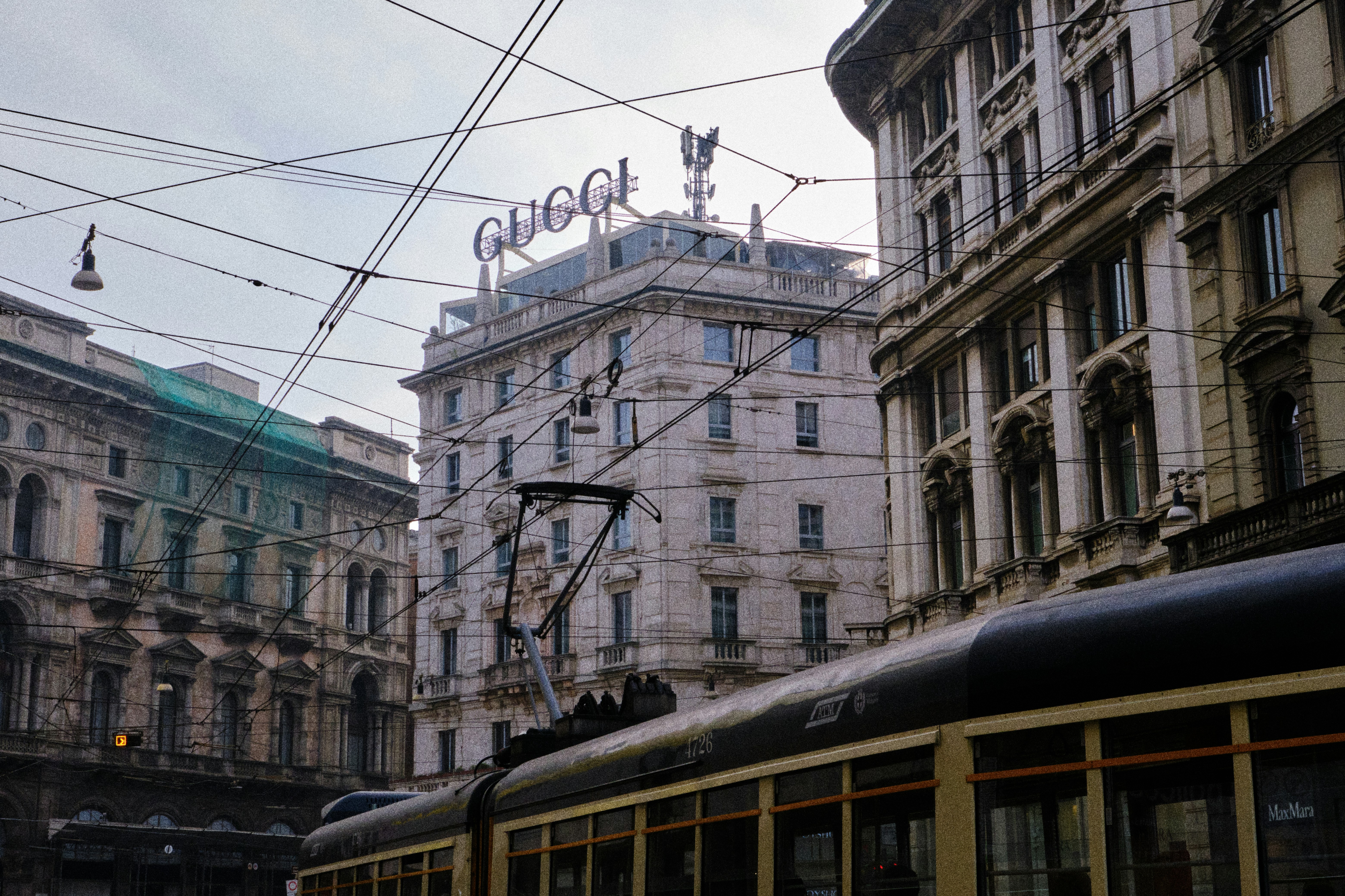 Tram passing by a historic building in Milan, showcasing the Gucci sign atop the structure. The scene captures the essence of urban life in a bustling city.