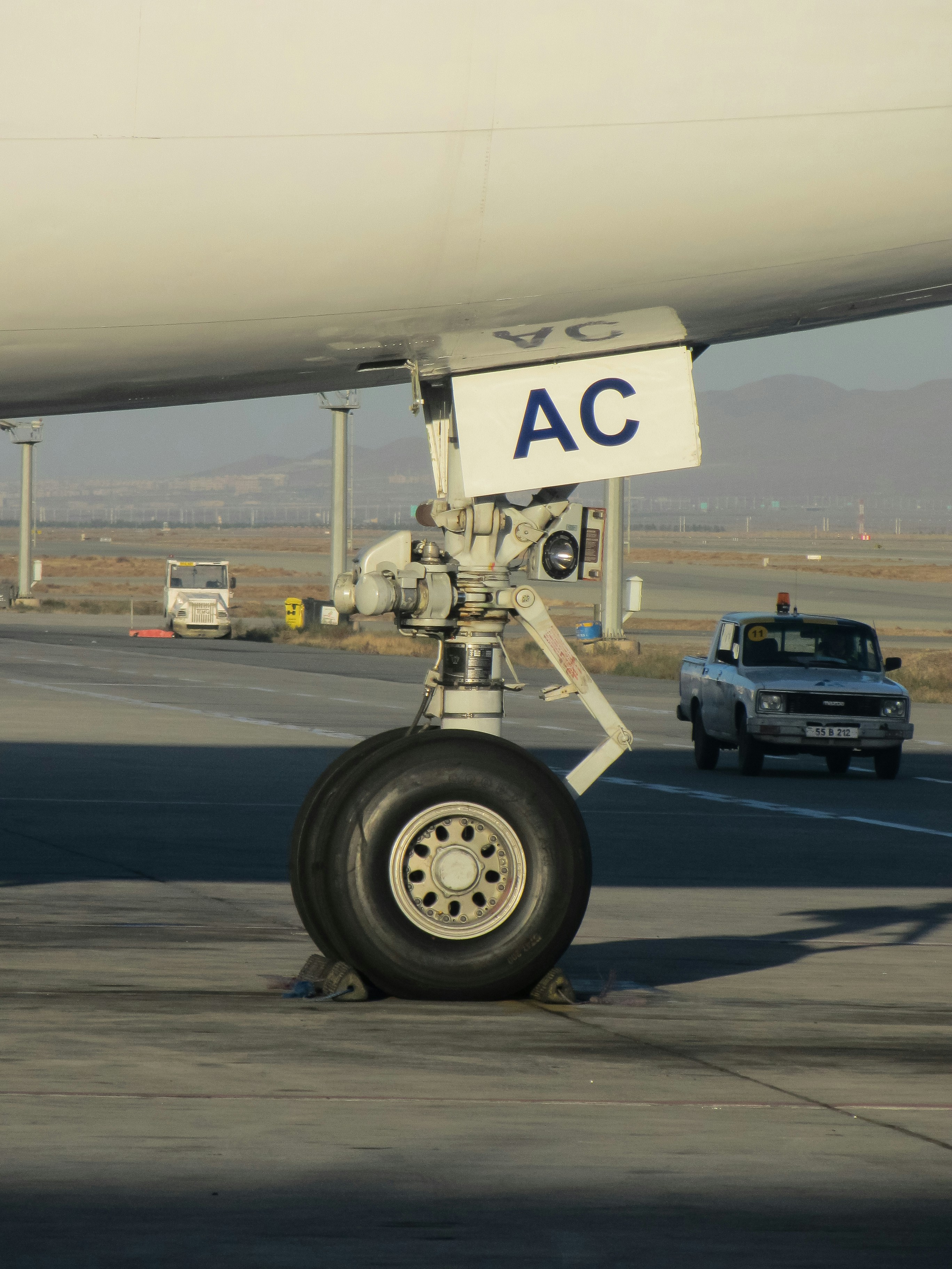 Close-up of a jet's landing gear on a sunlit airport apron, with a utility vehicle in the background.
