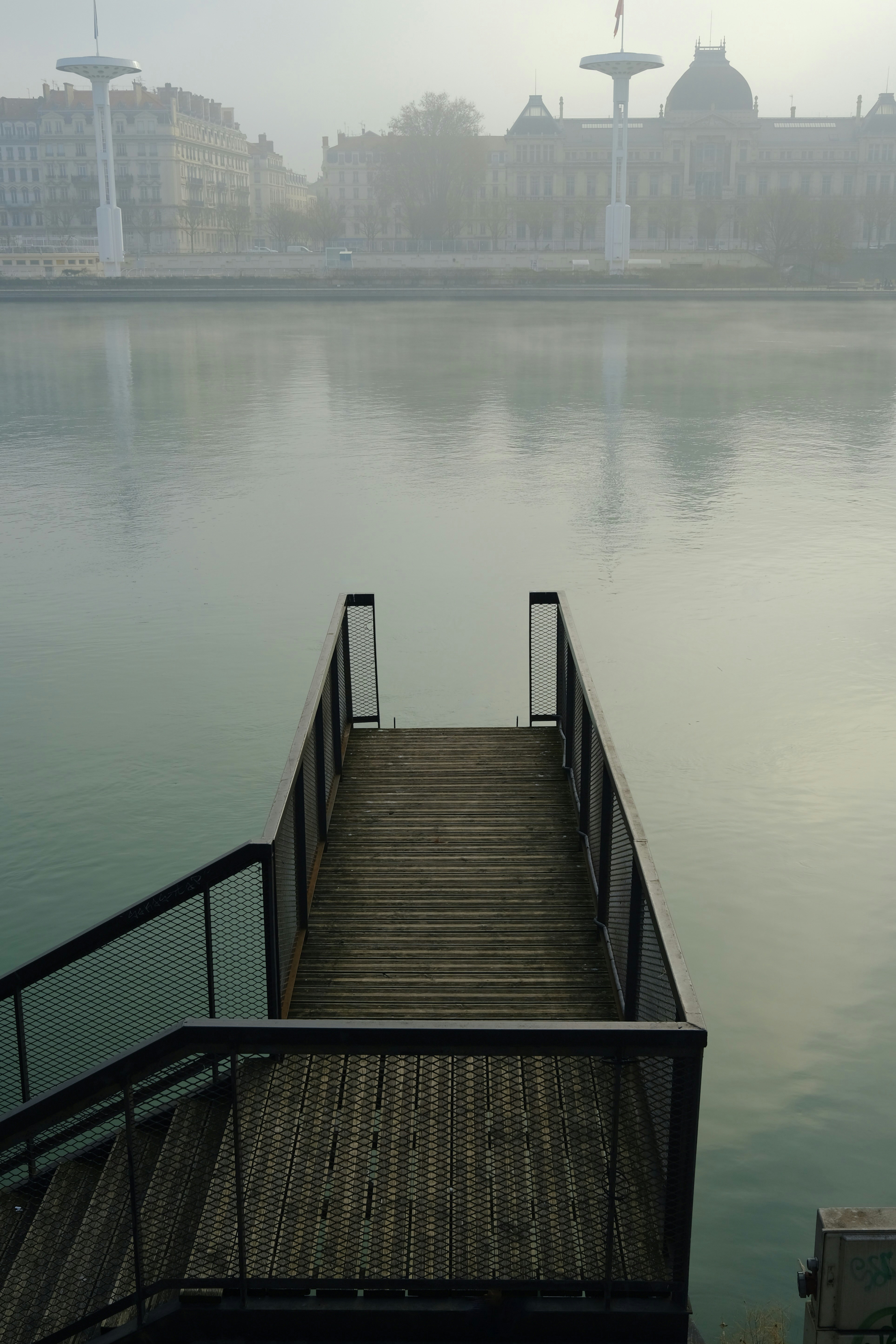 A serene dock stretches over calm waters, enveloped in a soft morning fog, with distant architectural silhouettes emerging through the haze.