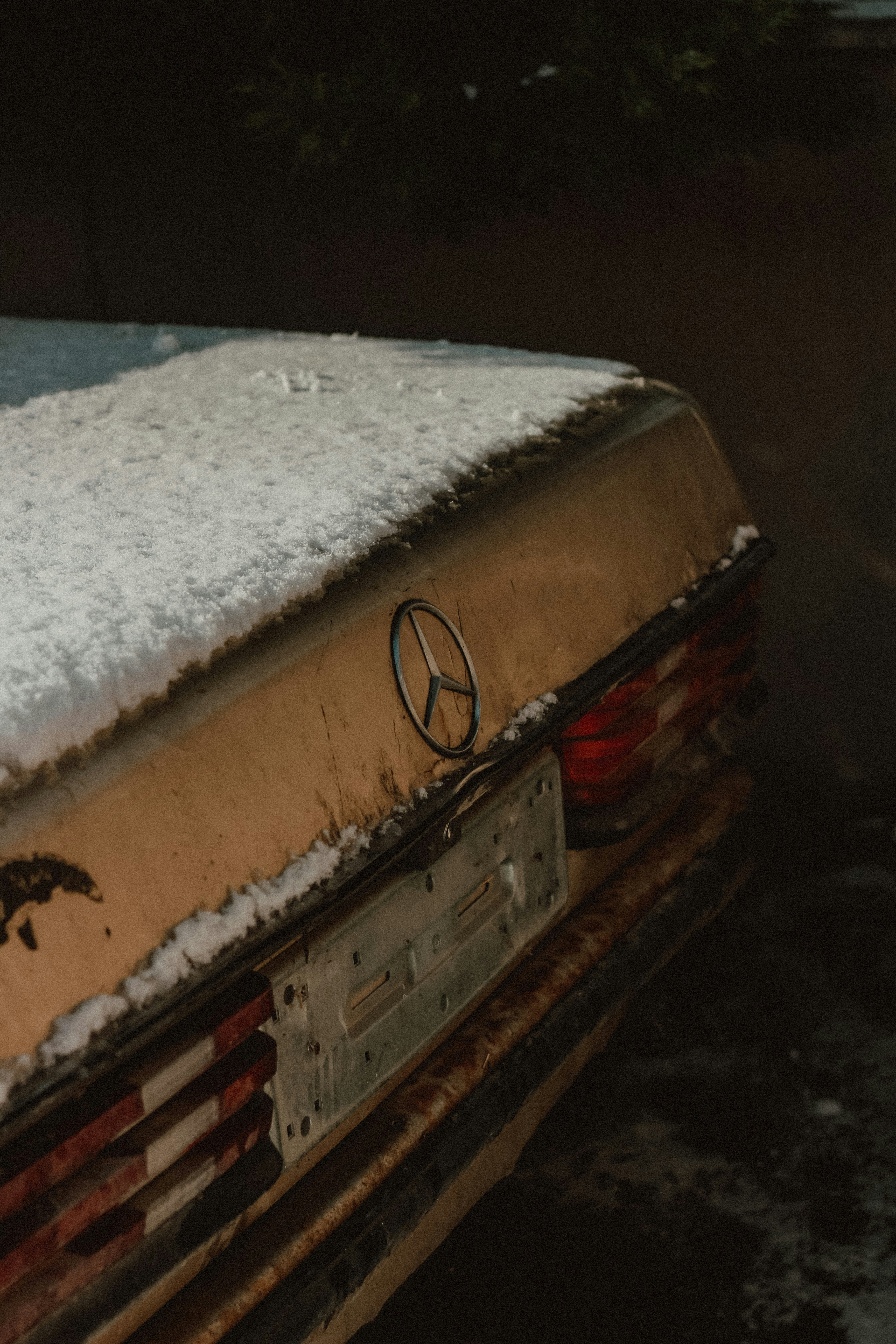 Snow-covered rear of a vintage Mercedes, showcasing its emblem and weathered paint. The scene evokes nostalgia and the passage of time.