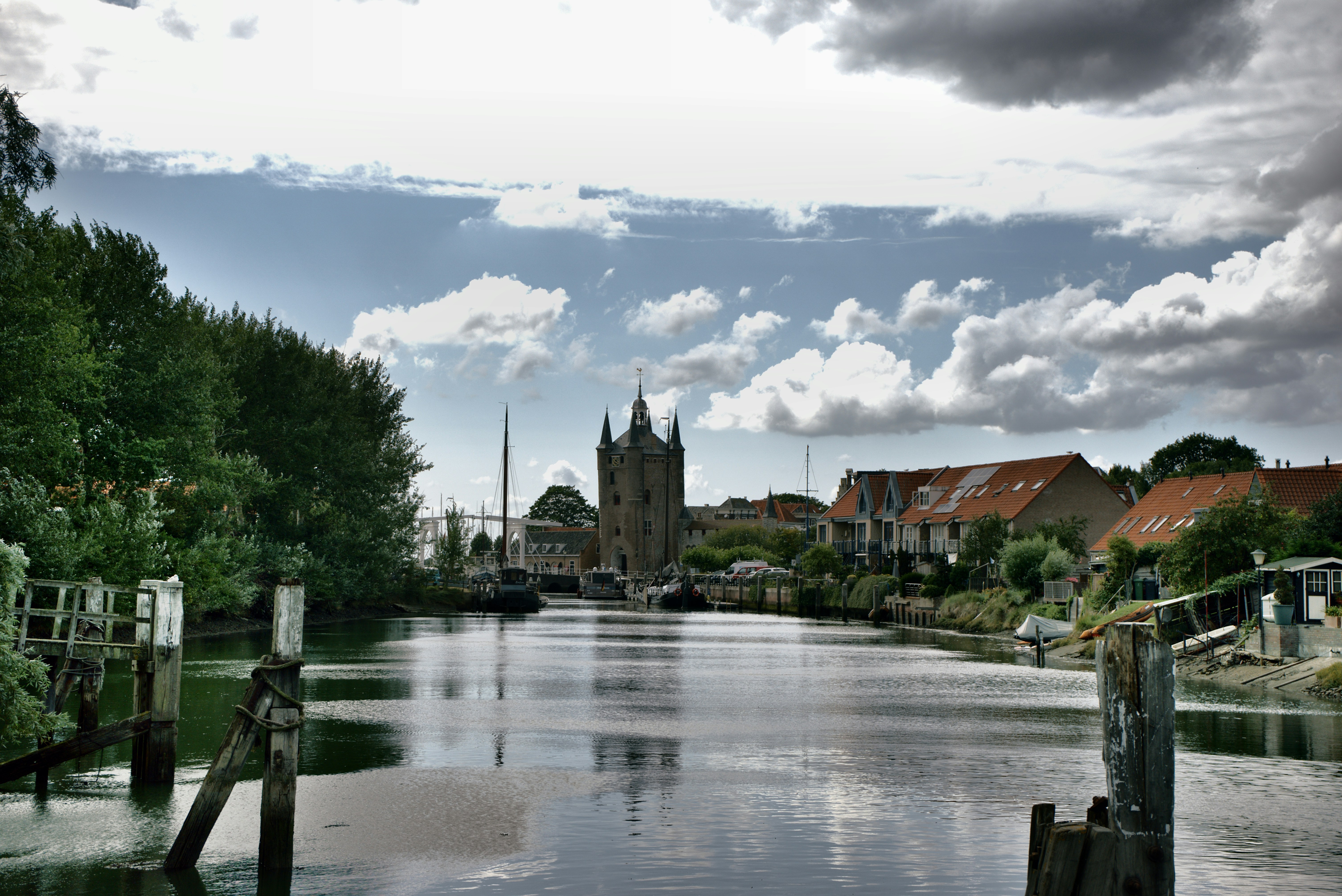 Historic tower overlooking a tranquil canal, framed by lush greenery and quaint buildings. Dramatic clouds add depth to the scene.