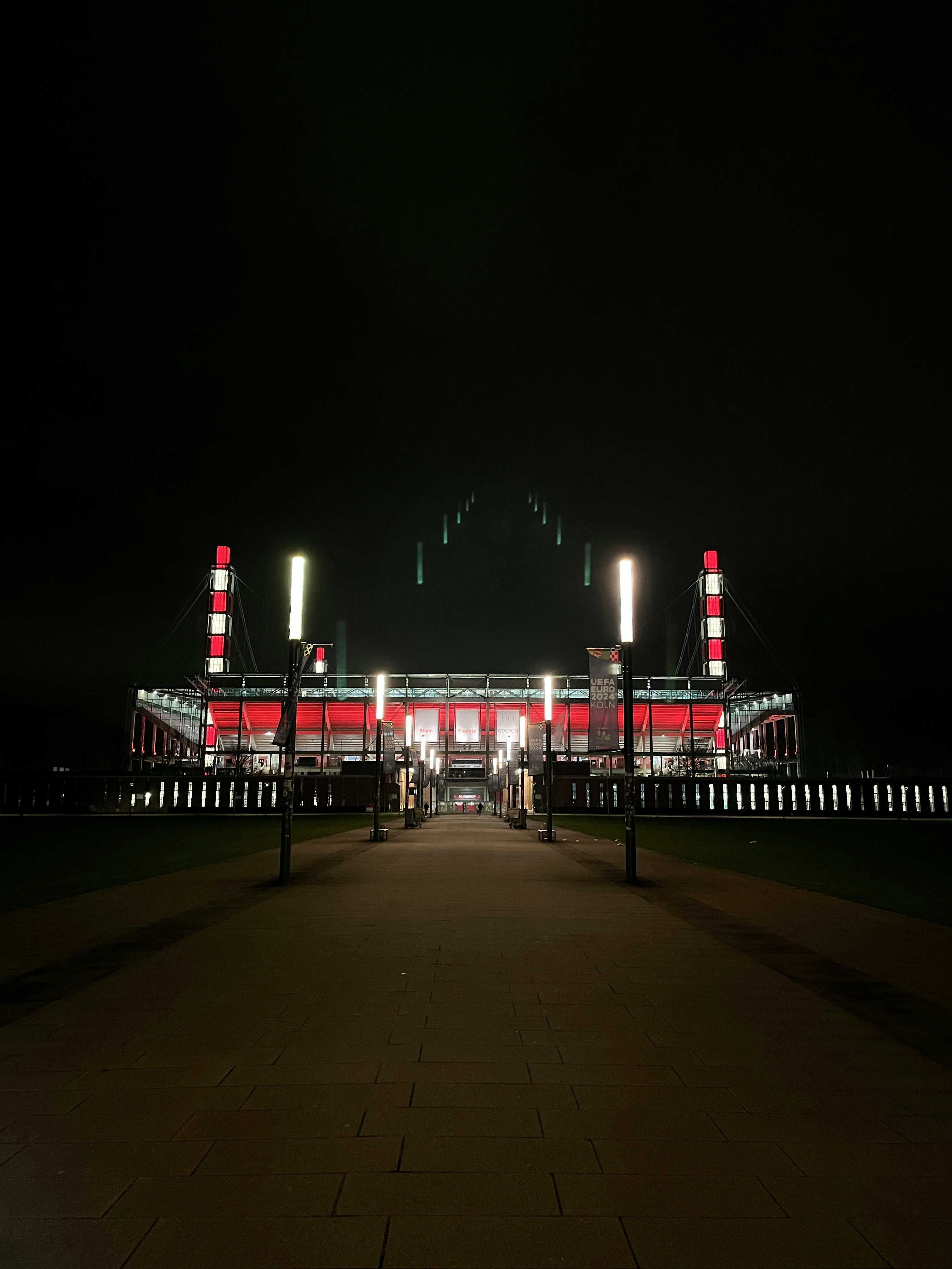 A vibrant sports stadium stands illuminated against a dark sky, with bright lights guiding the pathway leading to its entrance.