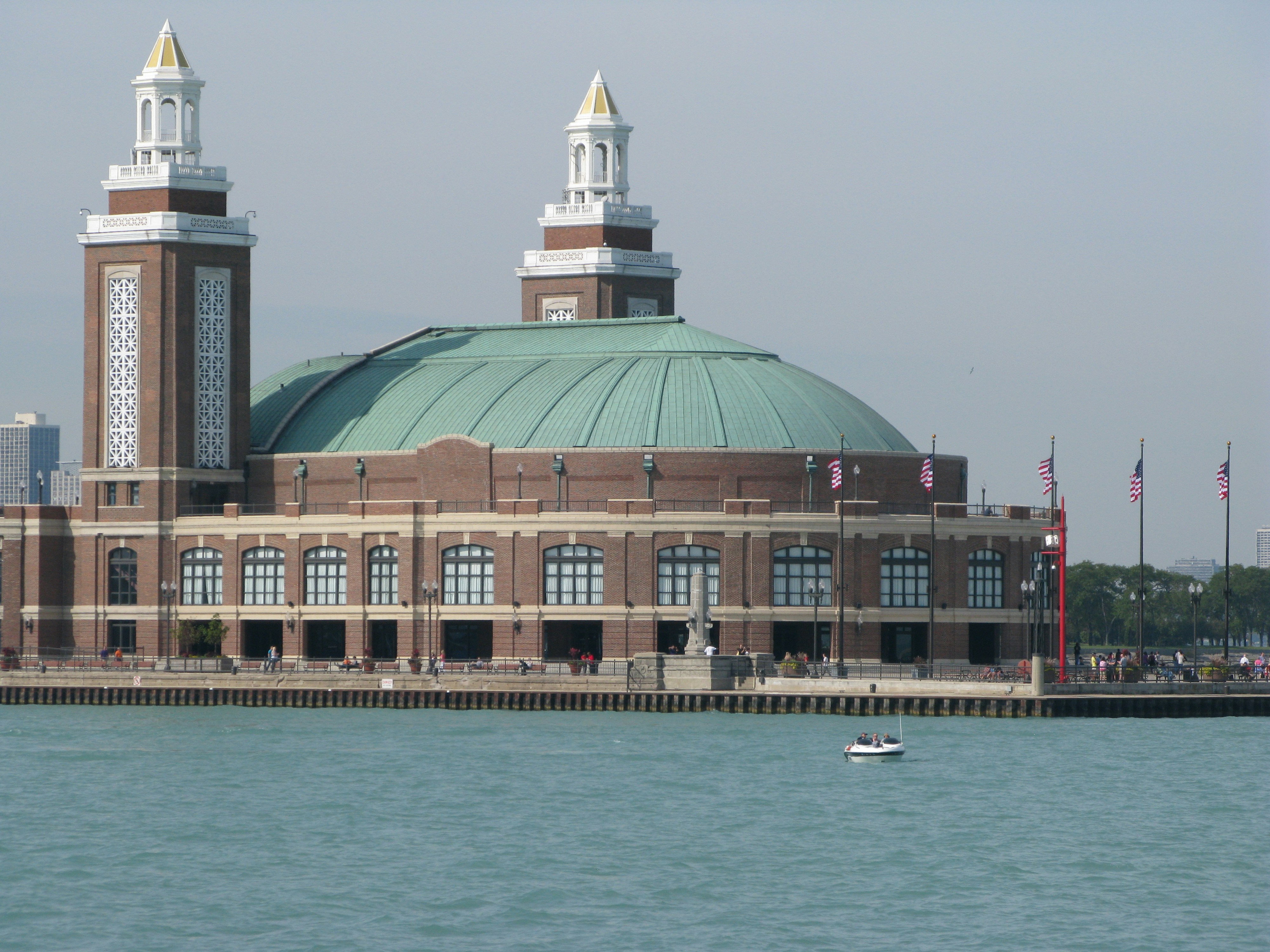 Historic waterfront structure with a distinctive green dome and twin towers, surrounded by flags and water. A small boat glides by, adding to the serene atmosphere.