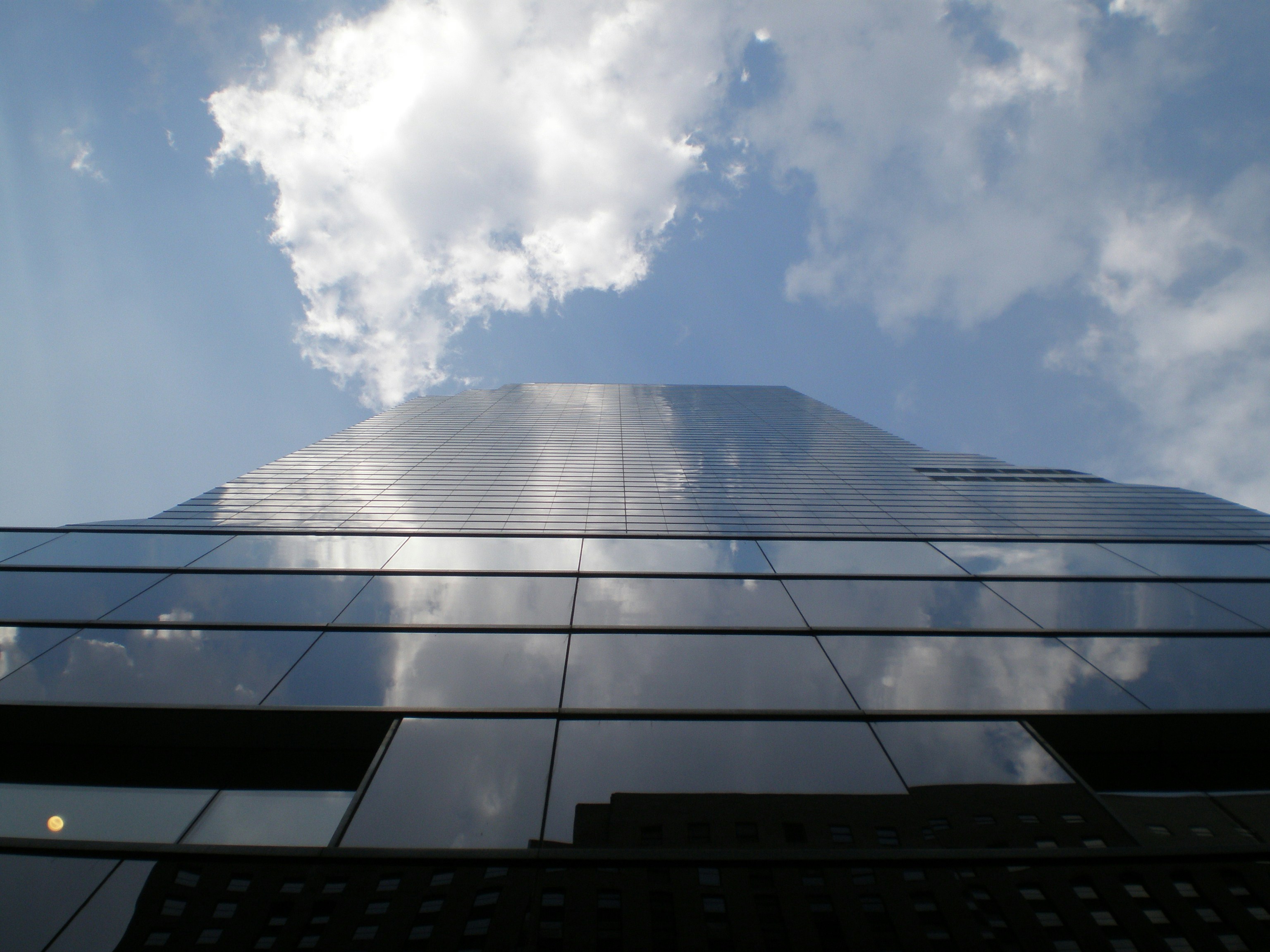 Photograph of a glass-clad skyscraper reflecting the blue sky and clouds, seen from ground level with vertical lines guiding the eye upward.