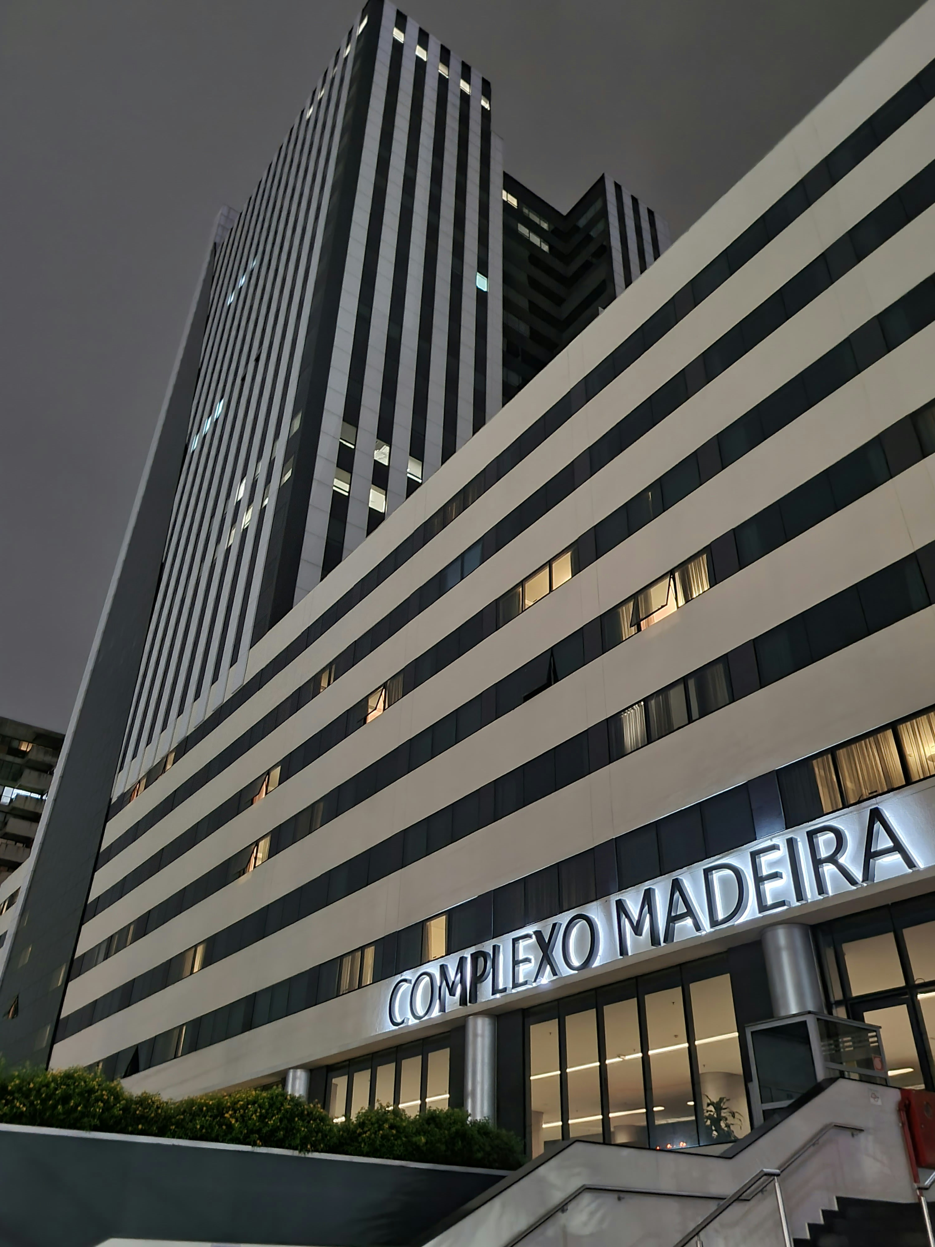 Modern high-rise building with illuminated windows and a clear sign reading 'Complexo Madeira' under a cloudy night sky.
