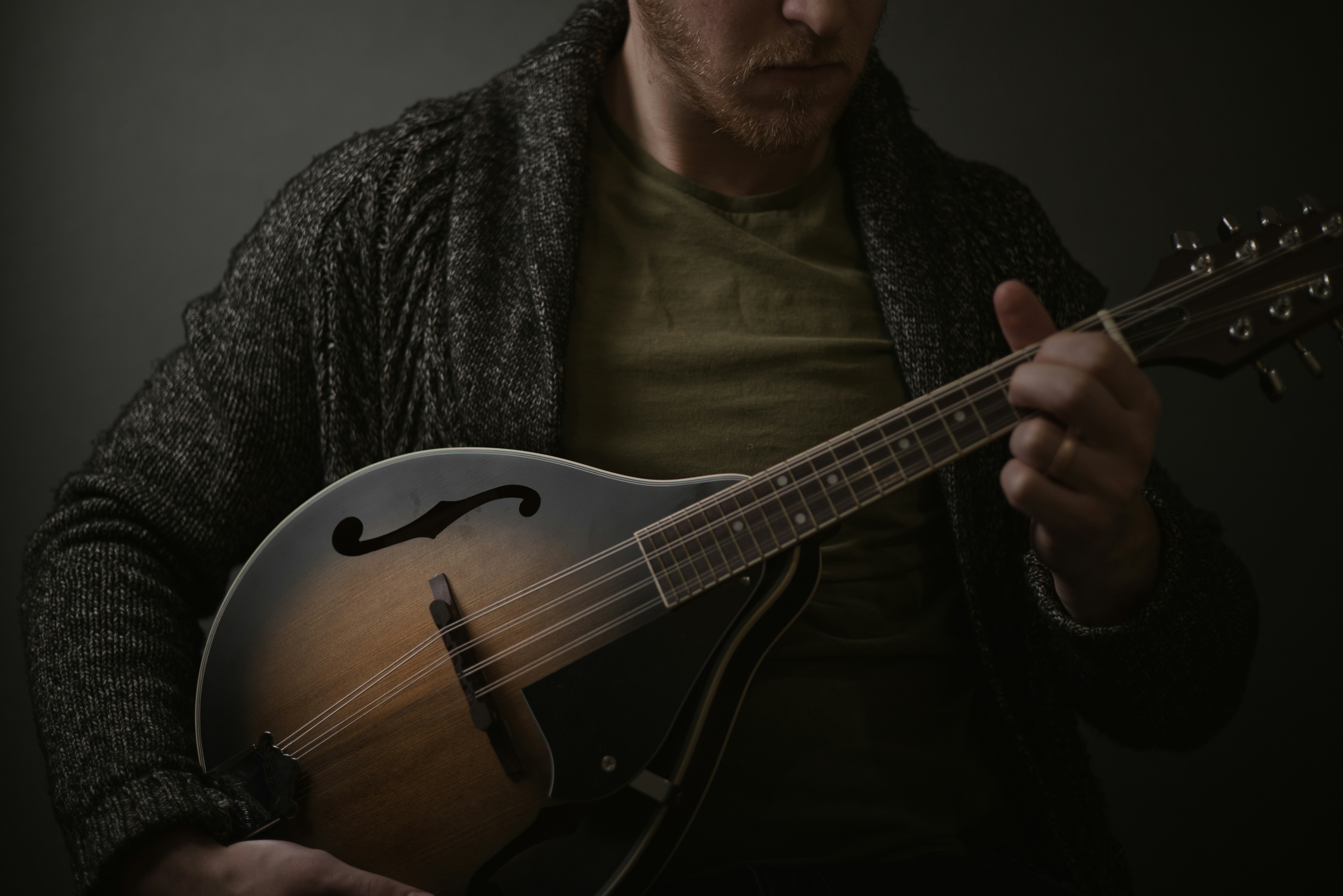 Muted lamplight reveals a guitarist cradling a mandolin, focusing on the instrument's curved body and the player's hands as they hover over the strings. A moody, intimate portrayal of a musician in twilight.