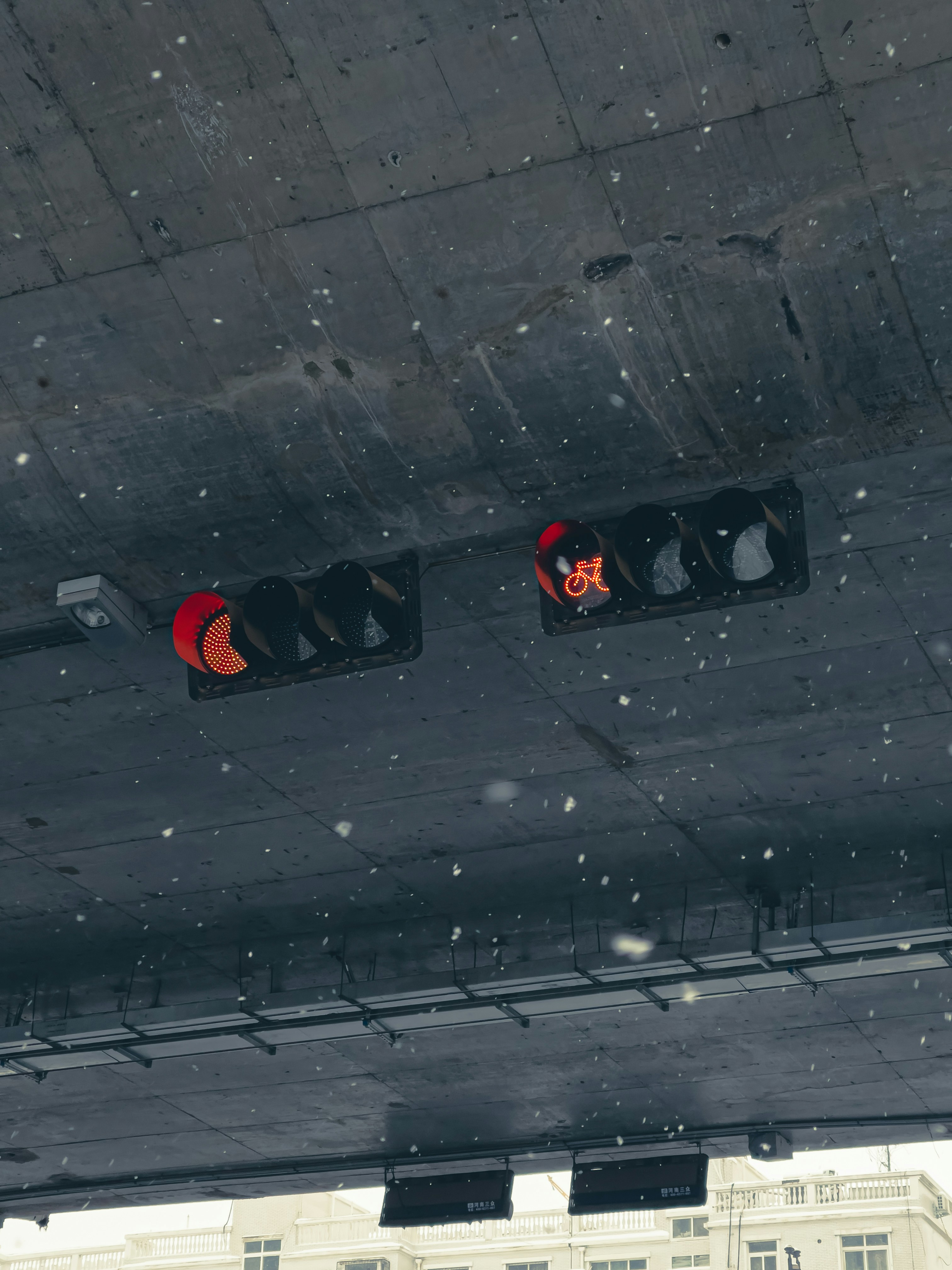 Traffic lights and a bicycle signal illuminated against a concrete ceiling, with snowflakes softly falling around them.
