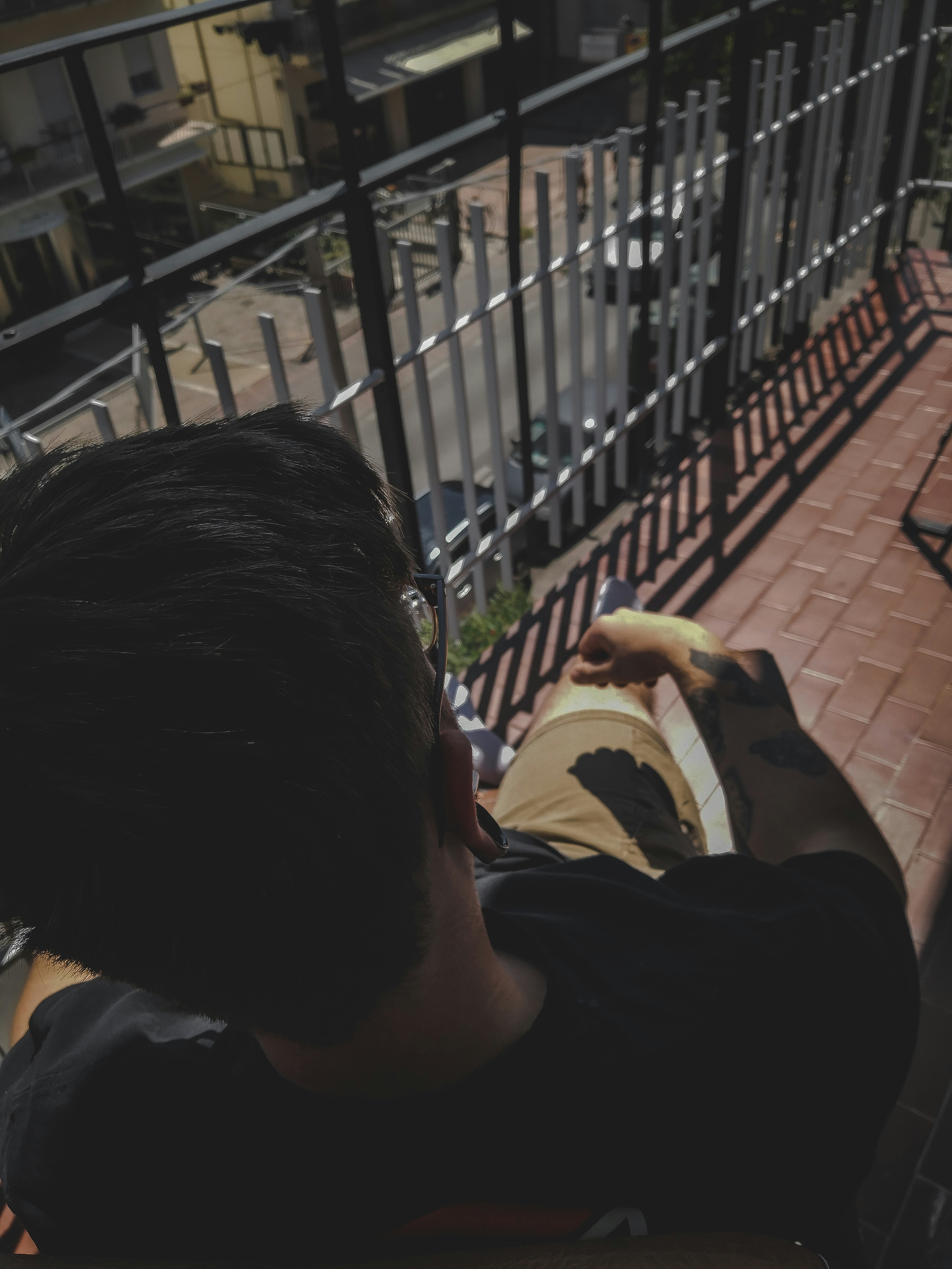 A person sitting on a balcony, enjoying a moment of reflection, framed by shadows and sunlight. The urban backdrop adds depth to the scene.