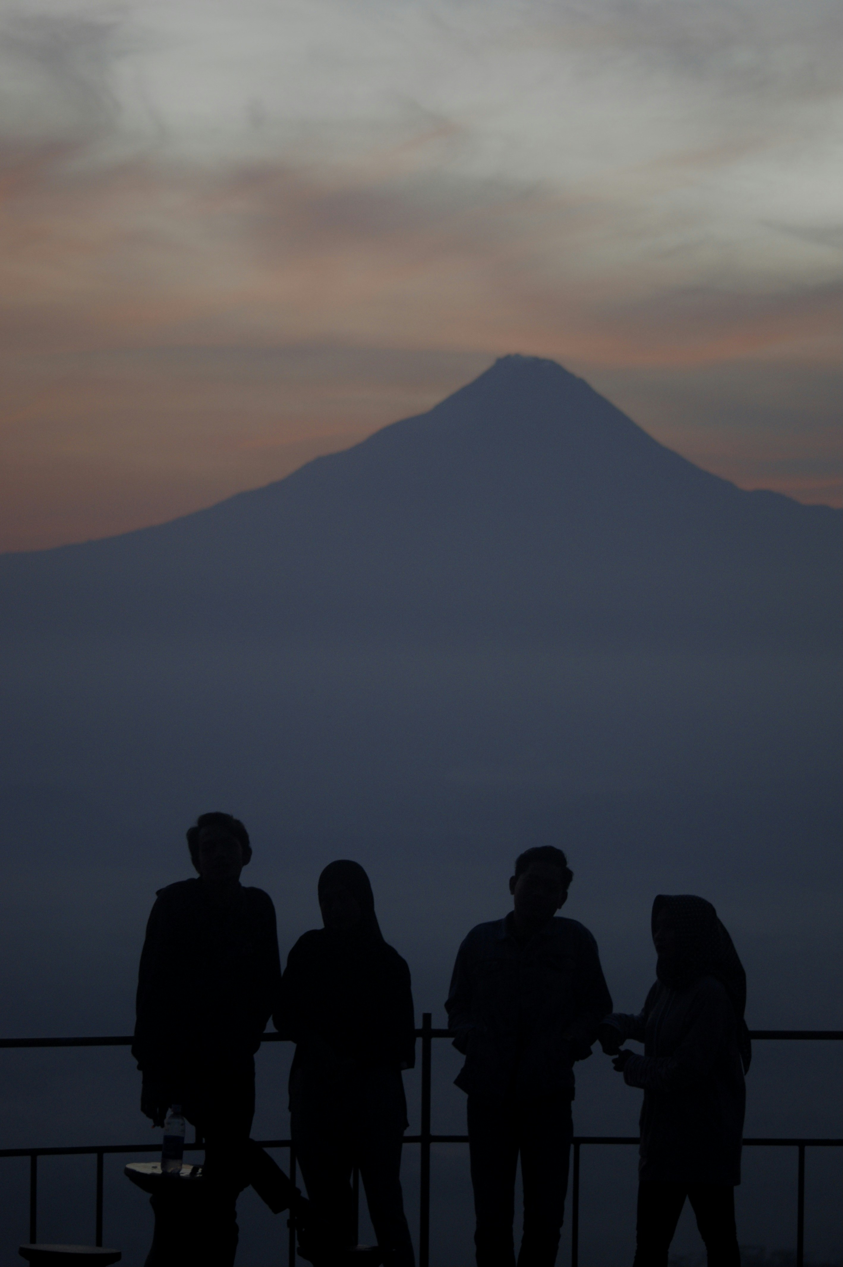 Silhouetted group stands along a railing at dusk as a distant volcano looms over a pastel sky. The scene emphasizes scale and mood in low light.