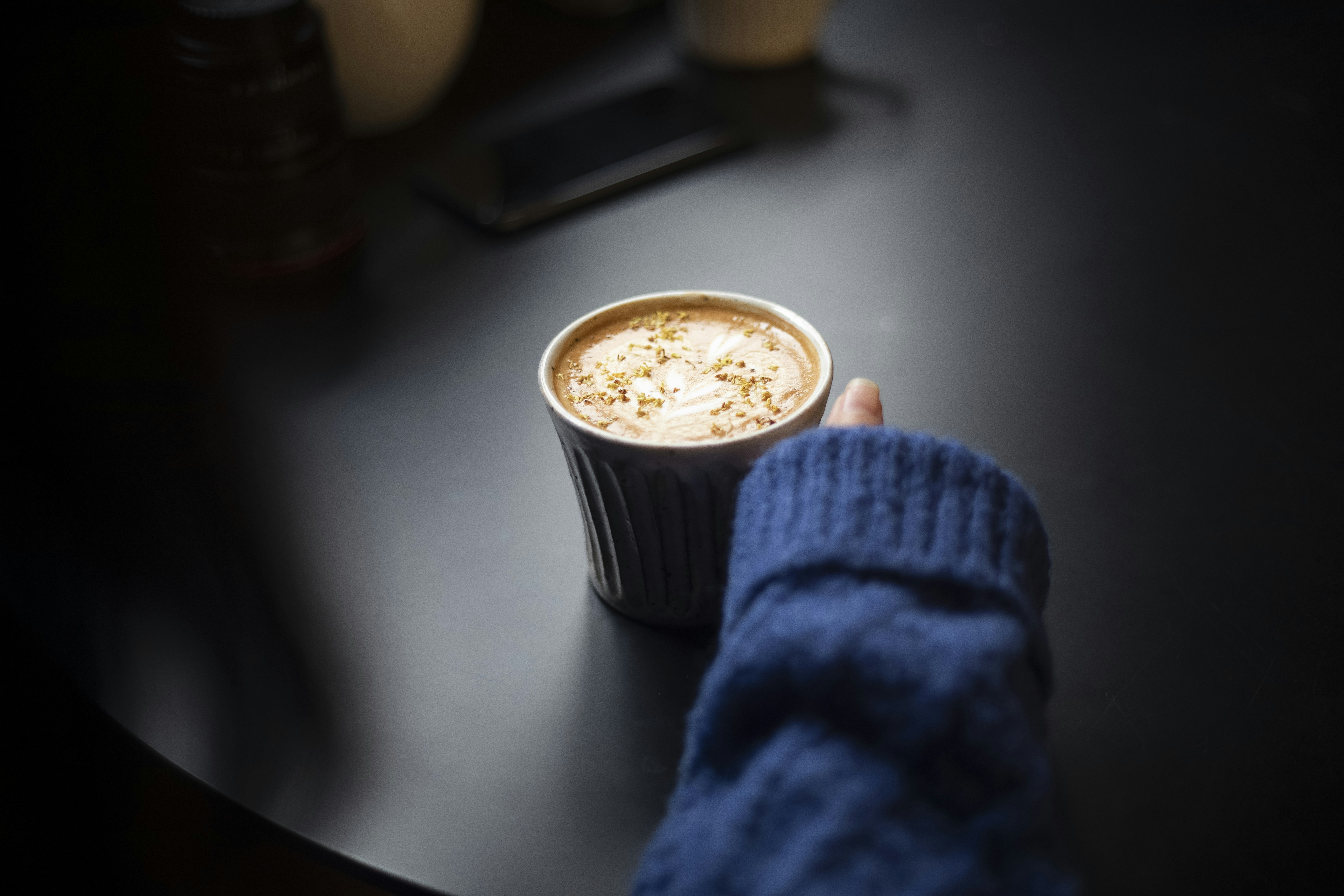A cozy hand in a blue sweater reaches for a cup of frothy beverage on a dark table, evoking a sense of warmth and relaxation.