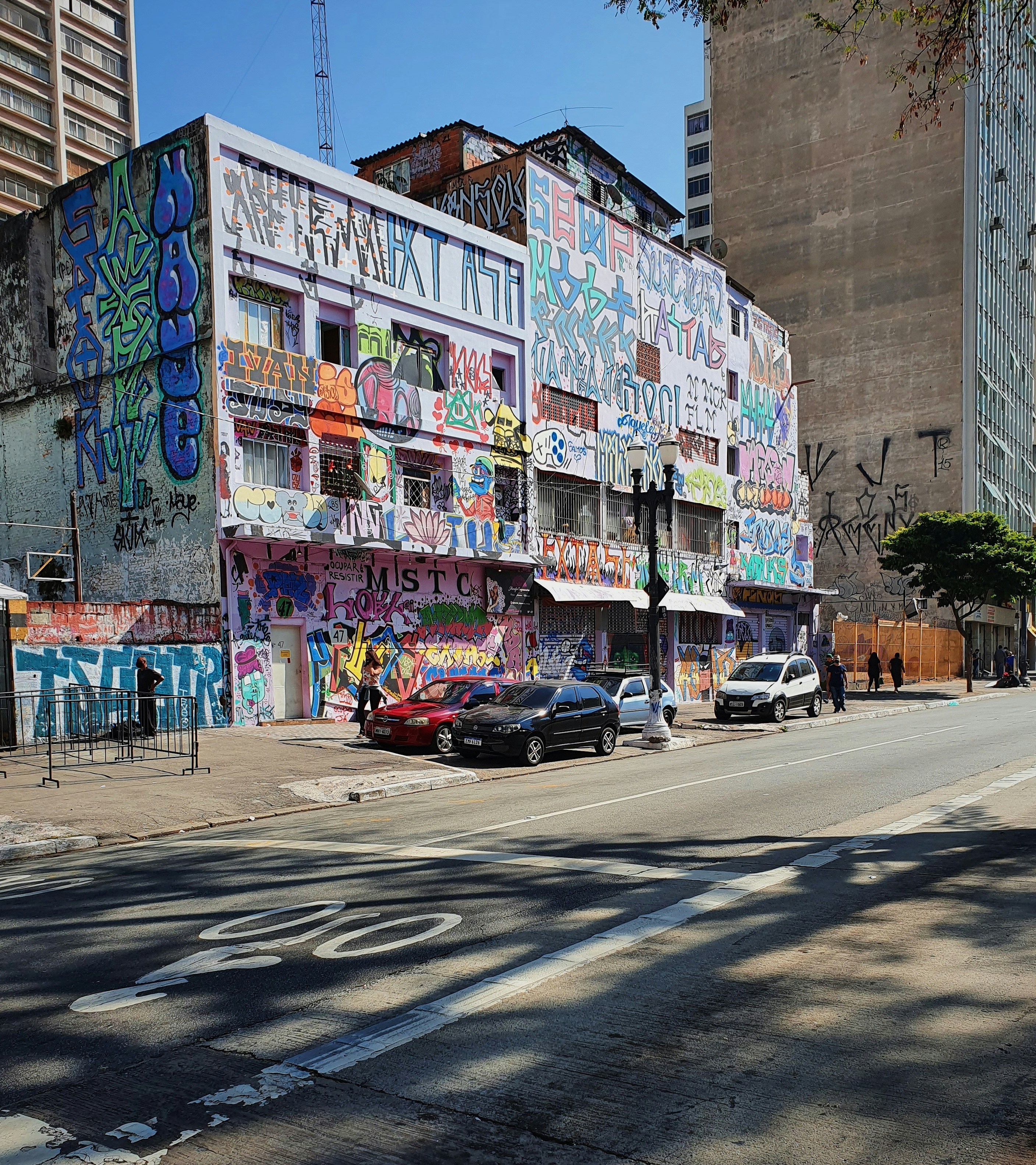 Vibrant street art adorns a building facade, showcasing a blend of colors and styles in an urban setting. Cars and pedestrians navigate the lively street scene.