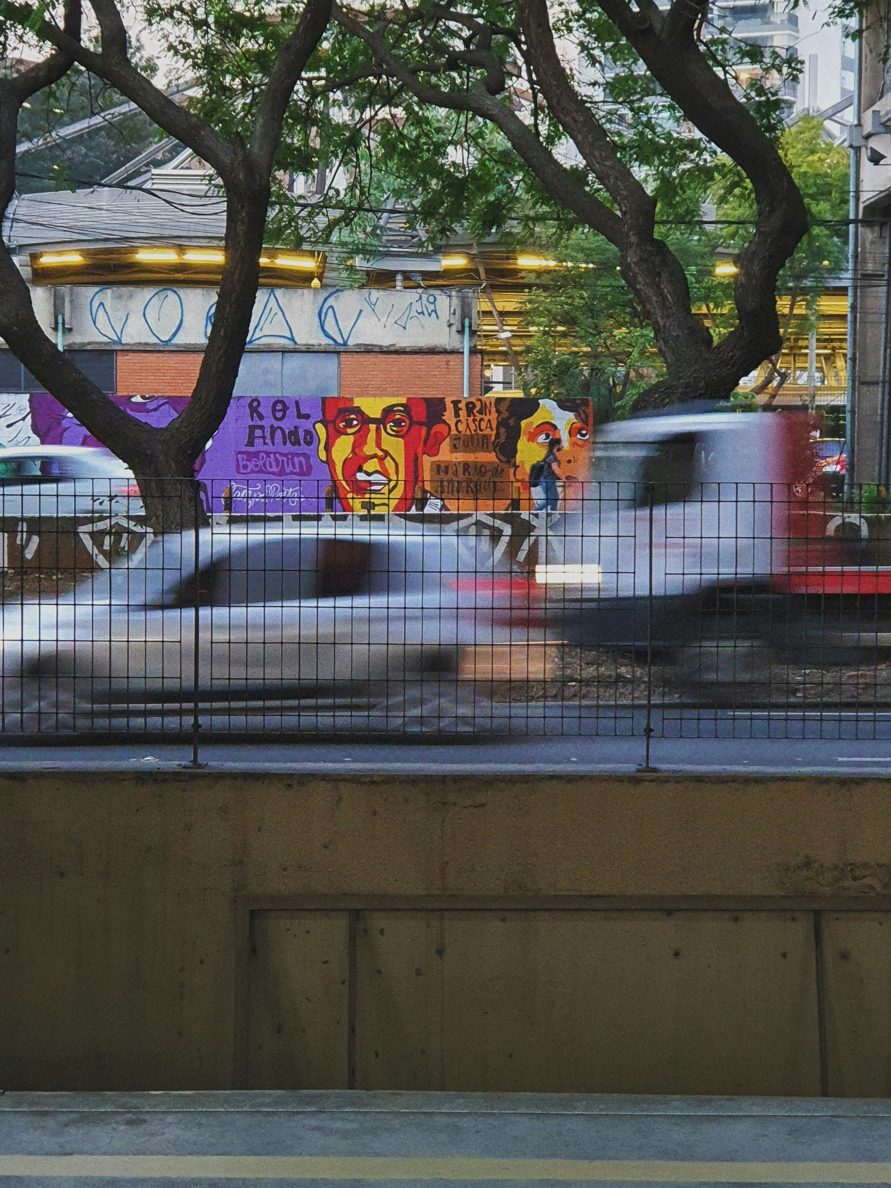 Vibrant street art featuring expressive faces on a wall, contrasted by the blurred motion of passing vehicles. The scene captures the dynamic energy of urban life.