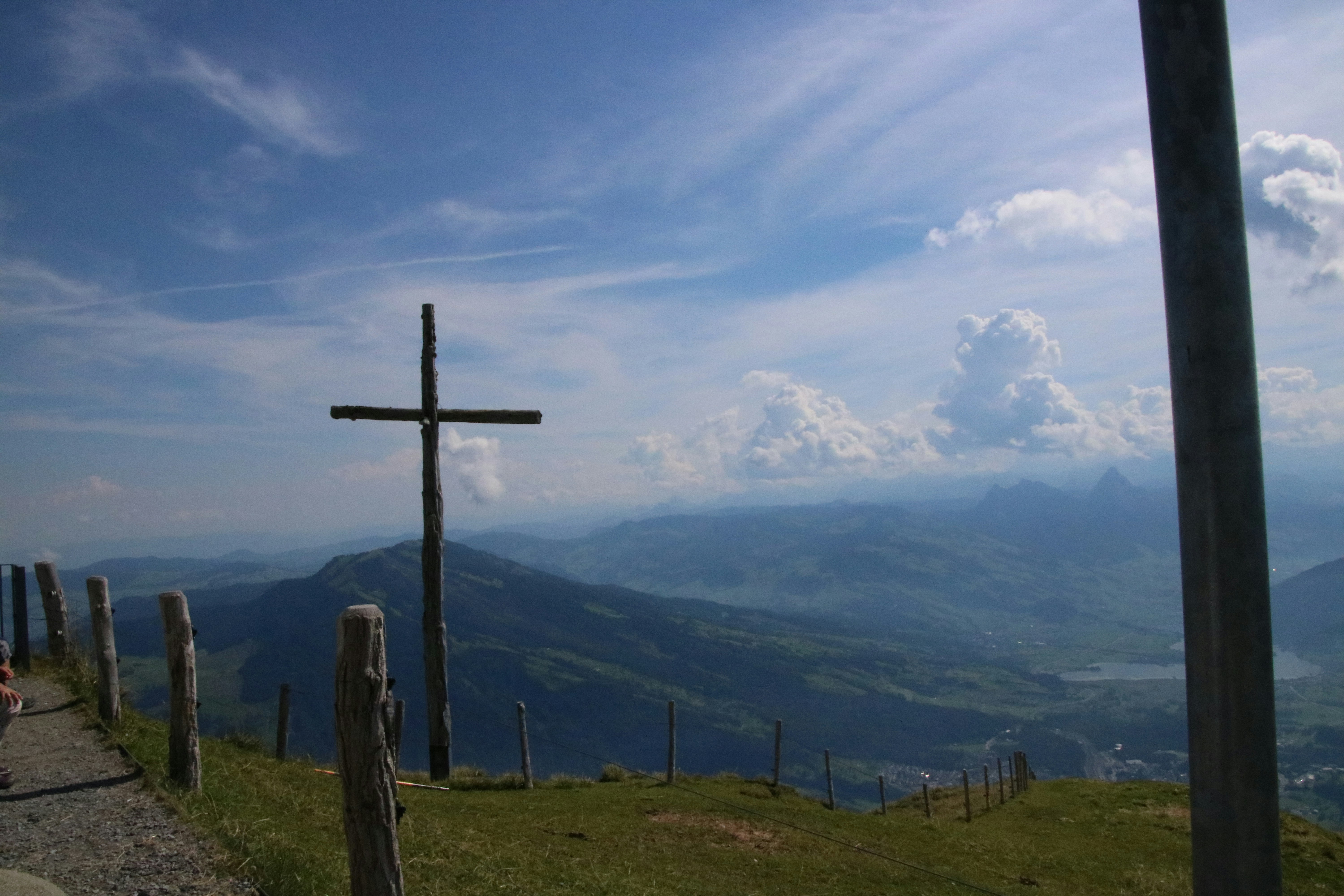 Wooden cross overlooking a vast mountainous landscape under a blue sky with fluffy clouds. The scene captures the serenity of nature at a high altitude.
