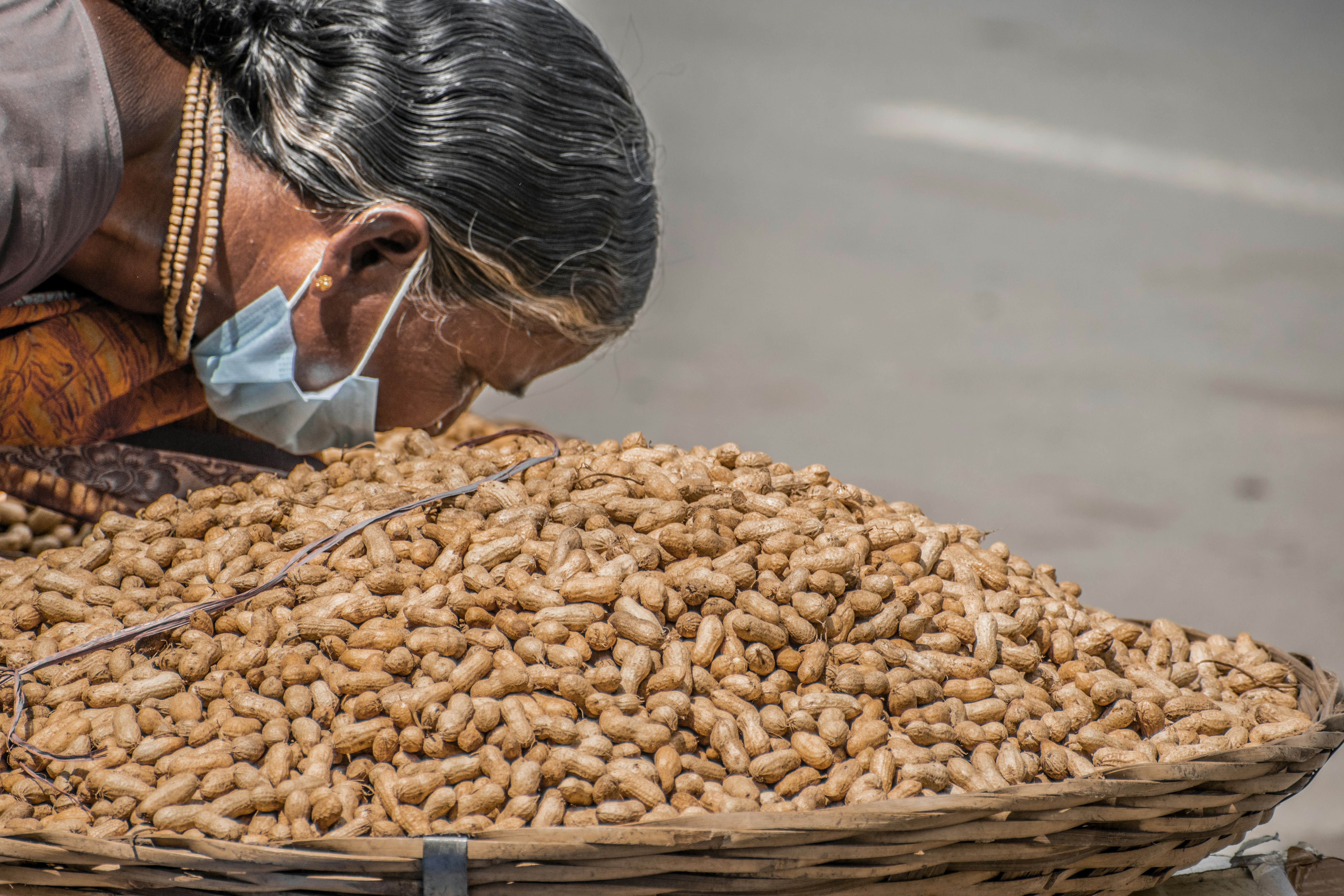 Woman wearing a face mask closely inspecting a basket full of peanuts on a street.