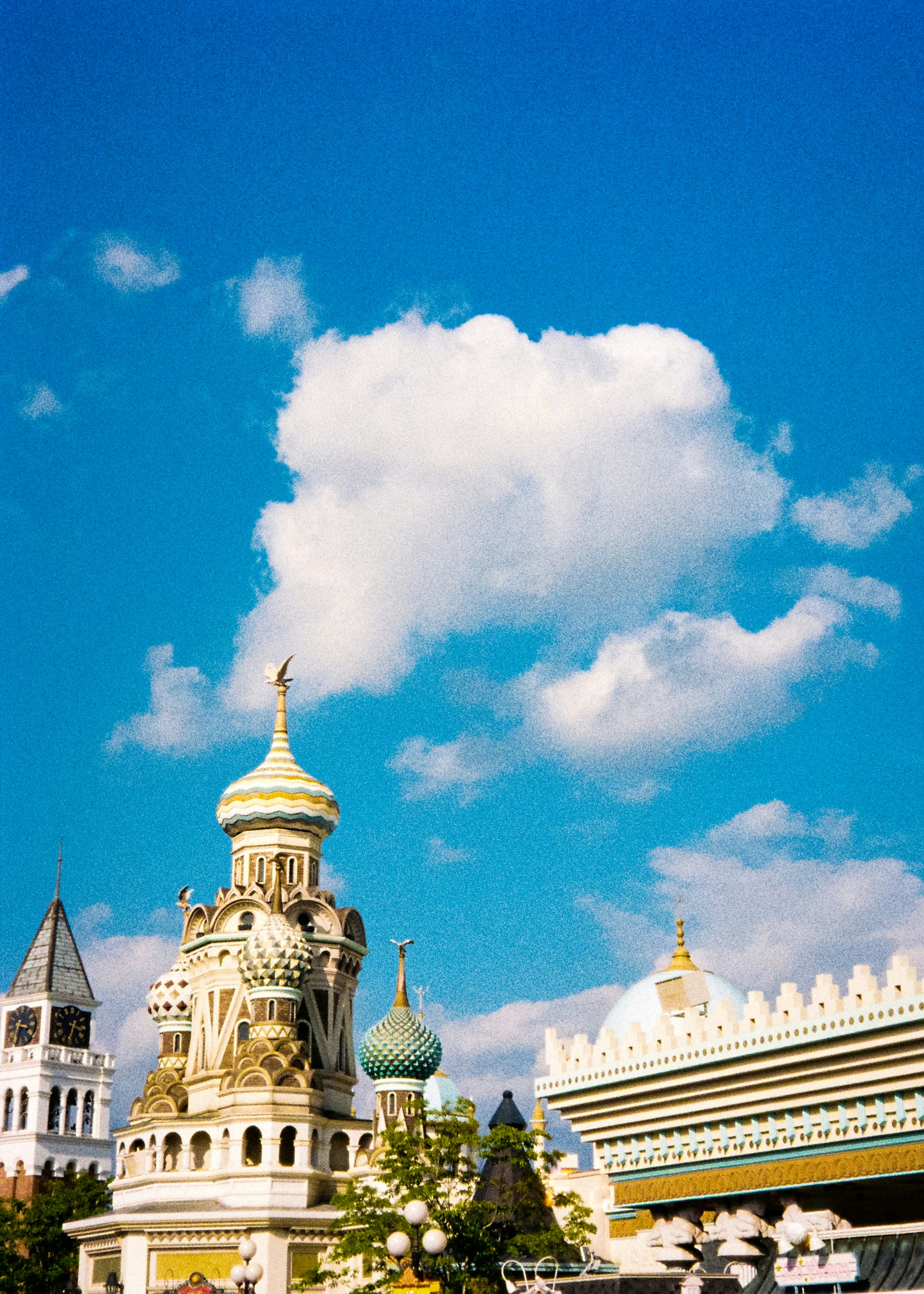 Intricate towers and domes rise against a vibrant blue sky, accented by fluffy clouds. The scene captures a blend of architectural styles and colors.