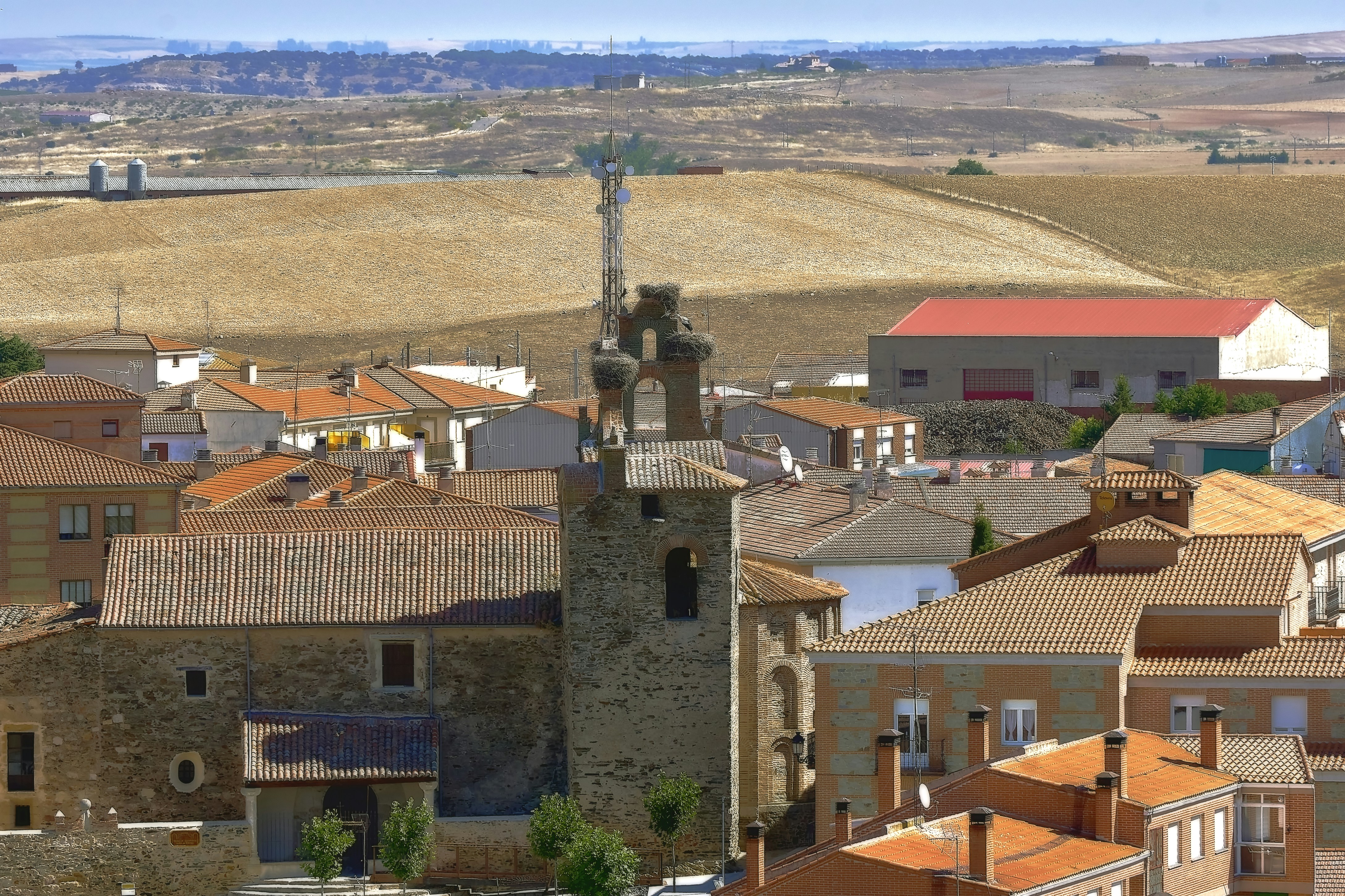 Clock tower rises above terracotta rooftops in a quaint village with expansive fields in the background.