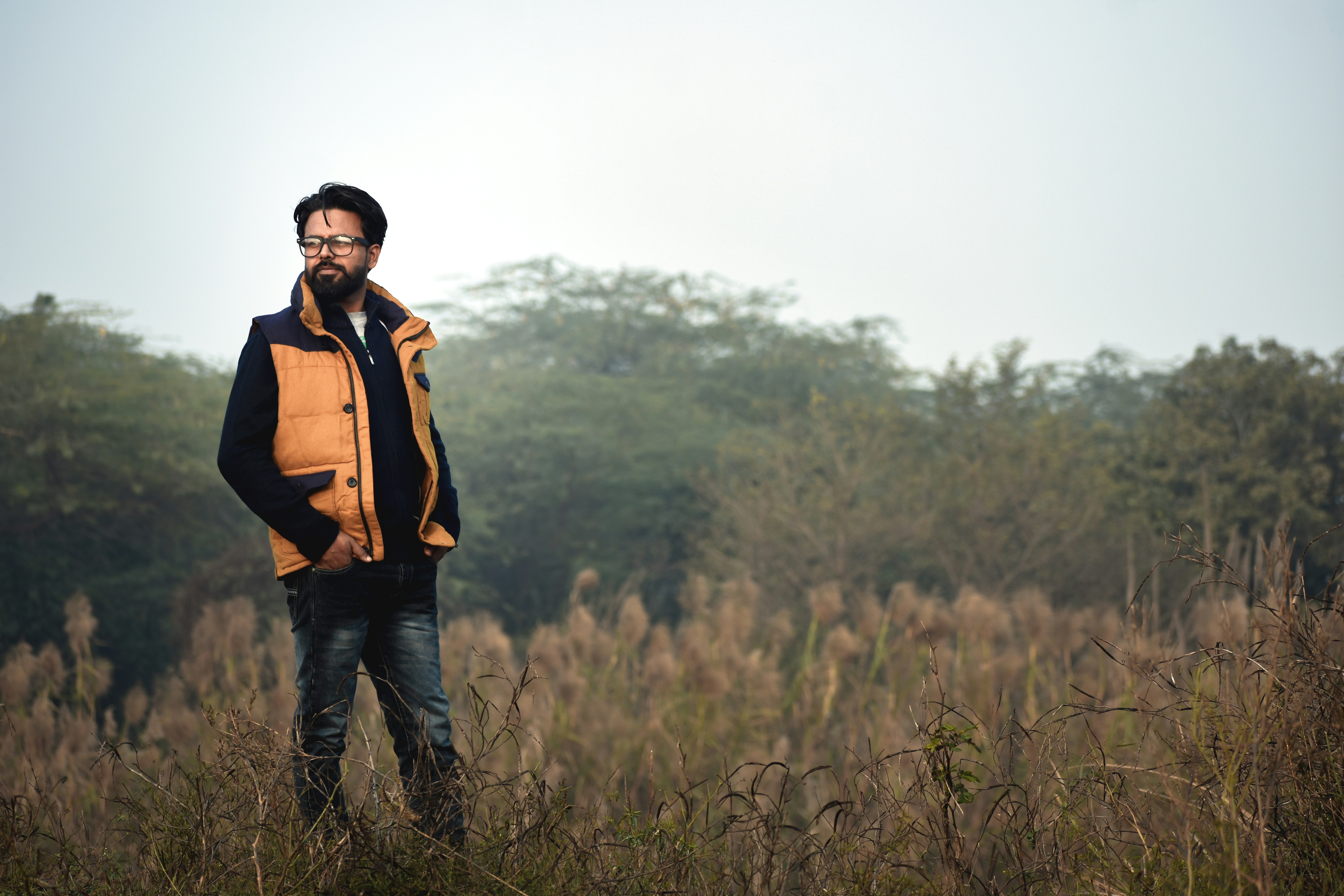 Person in a field wearing an orange vest, surrounded by tall grass and distant trees under a cloudy sky.