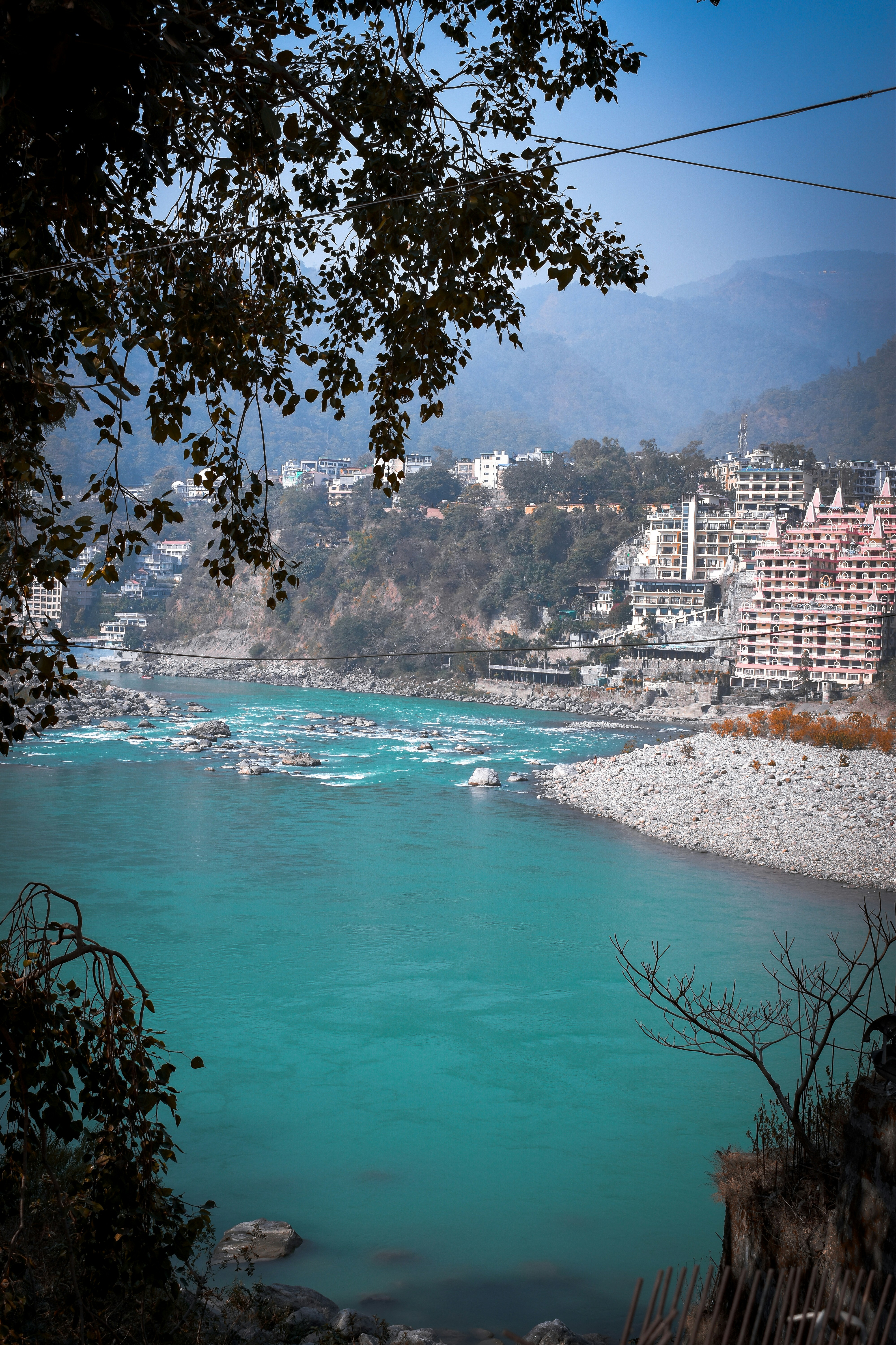 Turquoise river flowing gently past a bustling riverside town, framed by lush foliage and distant mountains.