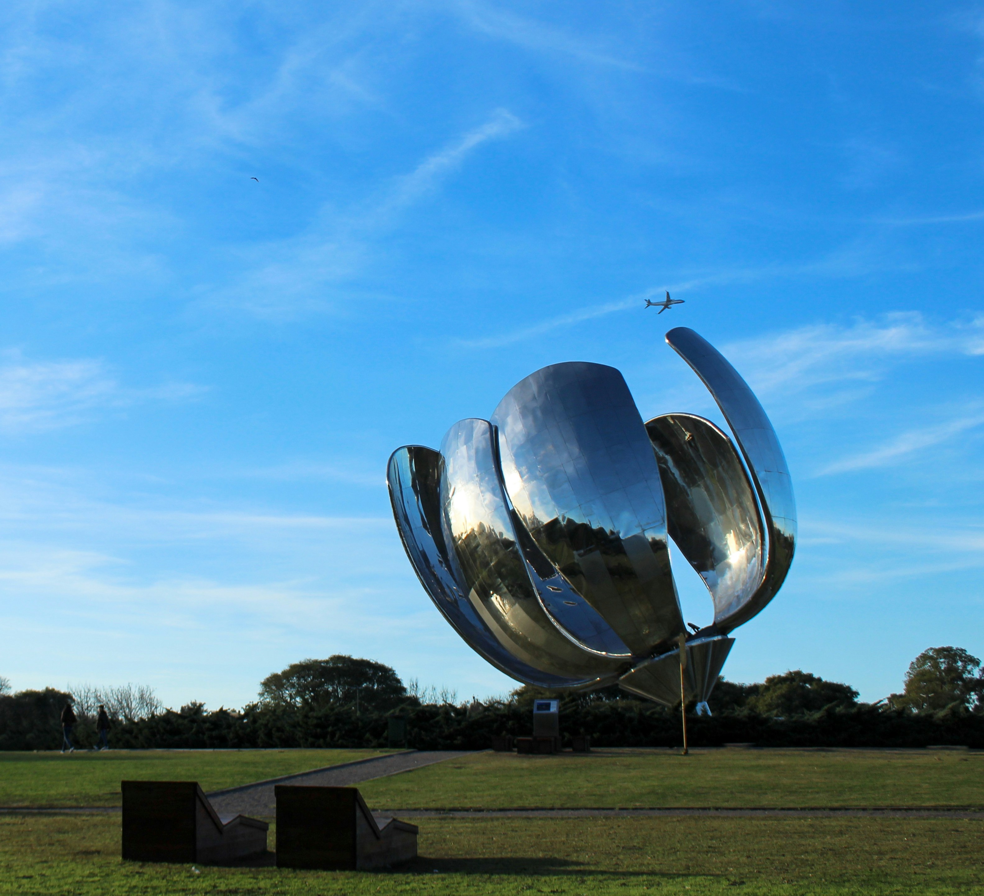A large, reflective floral sculpture stands prominently in a green field under a clear blue sky, with an airplane flying above.