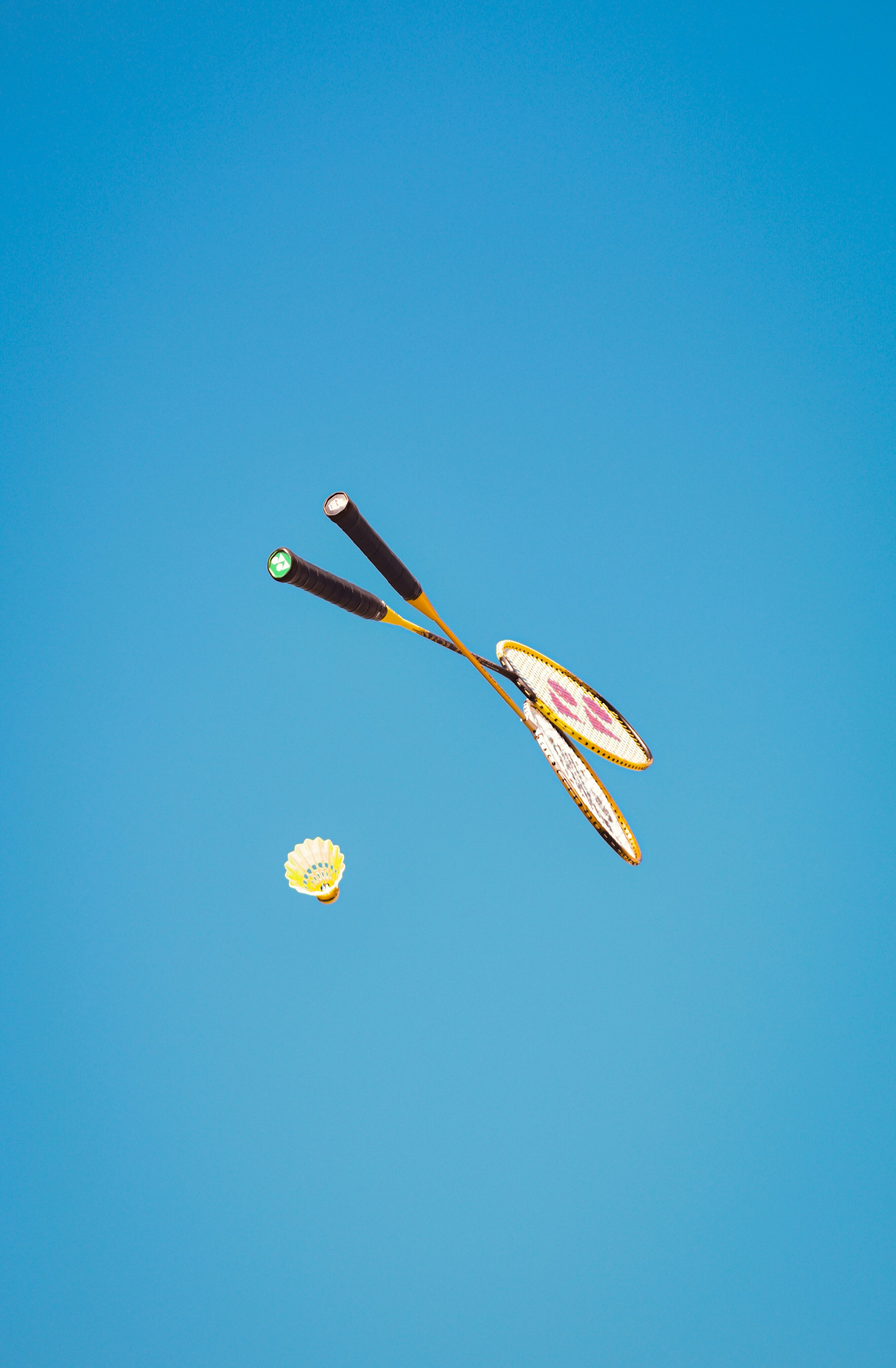 Two badminton rackets poised mid-air above a flying shuttlecock against a clear blue sky.