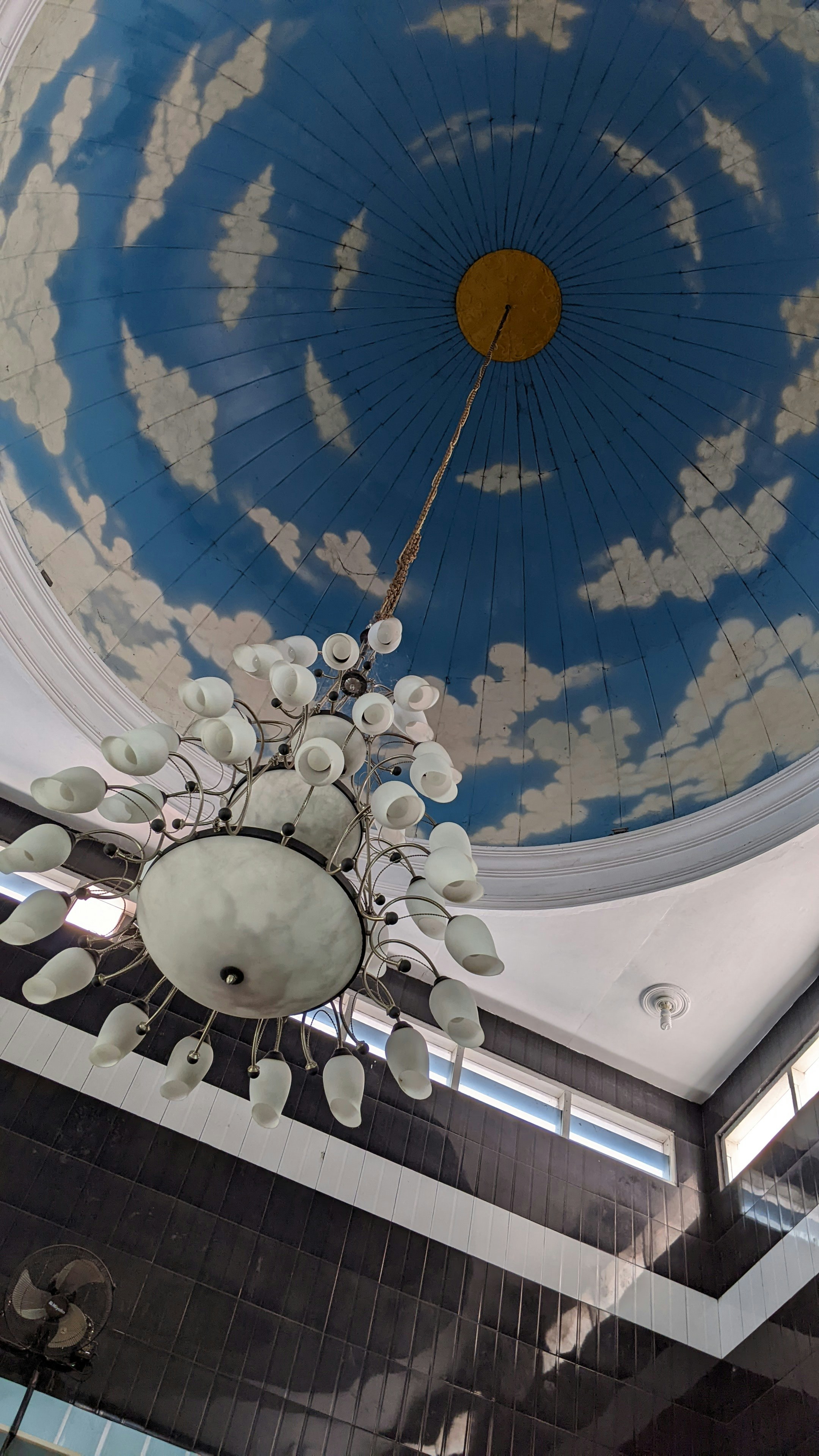 Blue sky dome with a central yellow disk and radiating lines, foregrounded by a circular chandelier with multiple white bulbs. Architectural photography highlighting the contrast between celestial ceiling and urban interior hardware.
