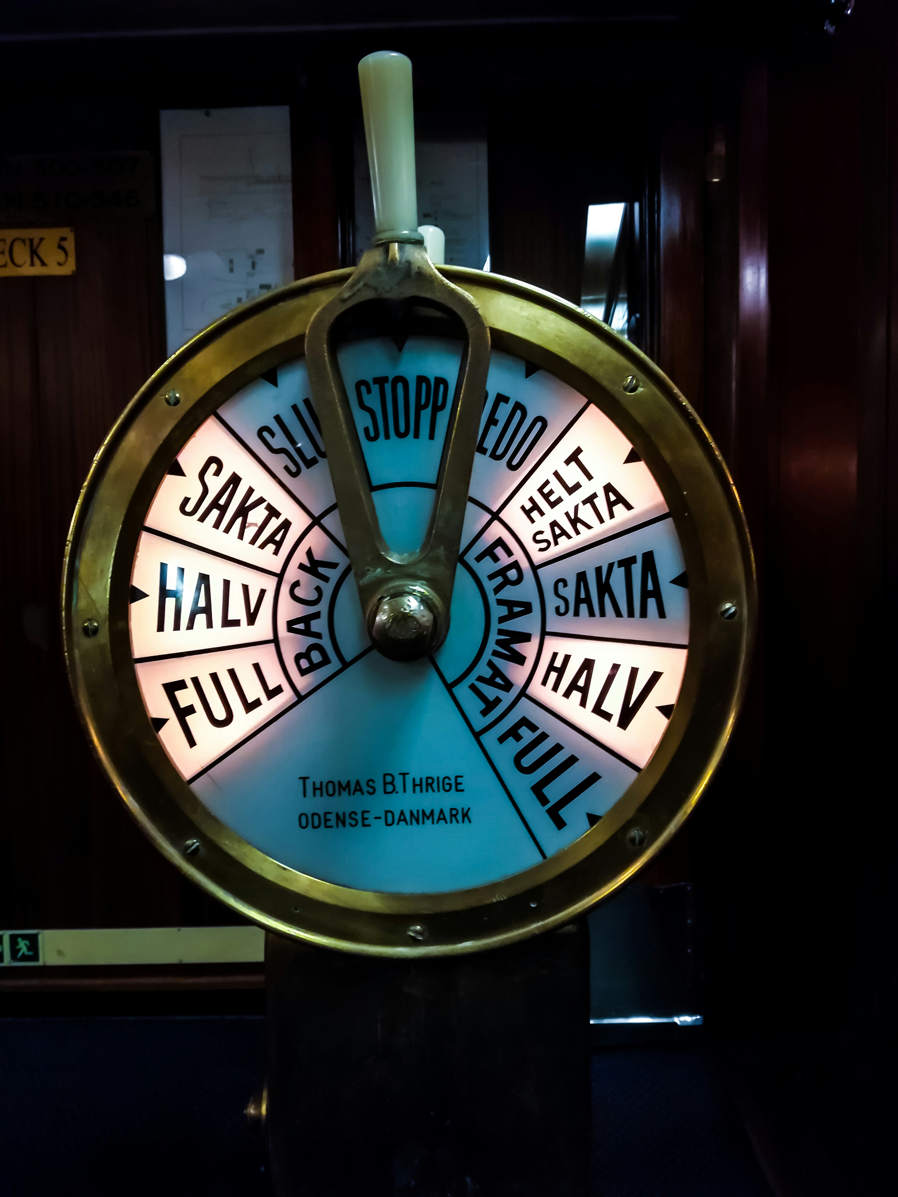 A vintage brass ship wheel with a blue-and-white segmented dial sits on a dark wooden deck, illuminated to emphasize its circular face and central spokes.